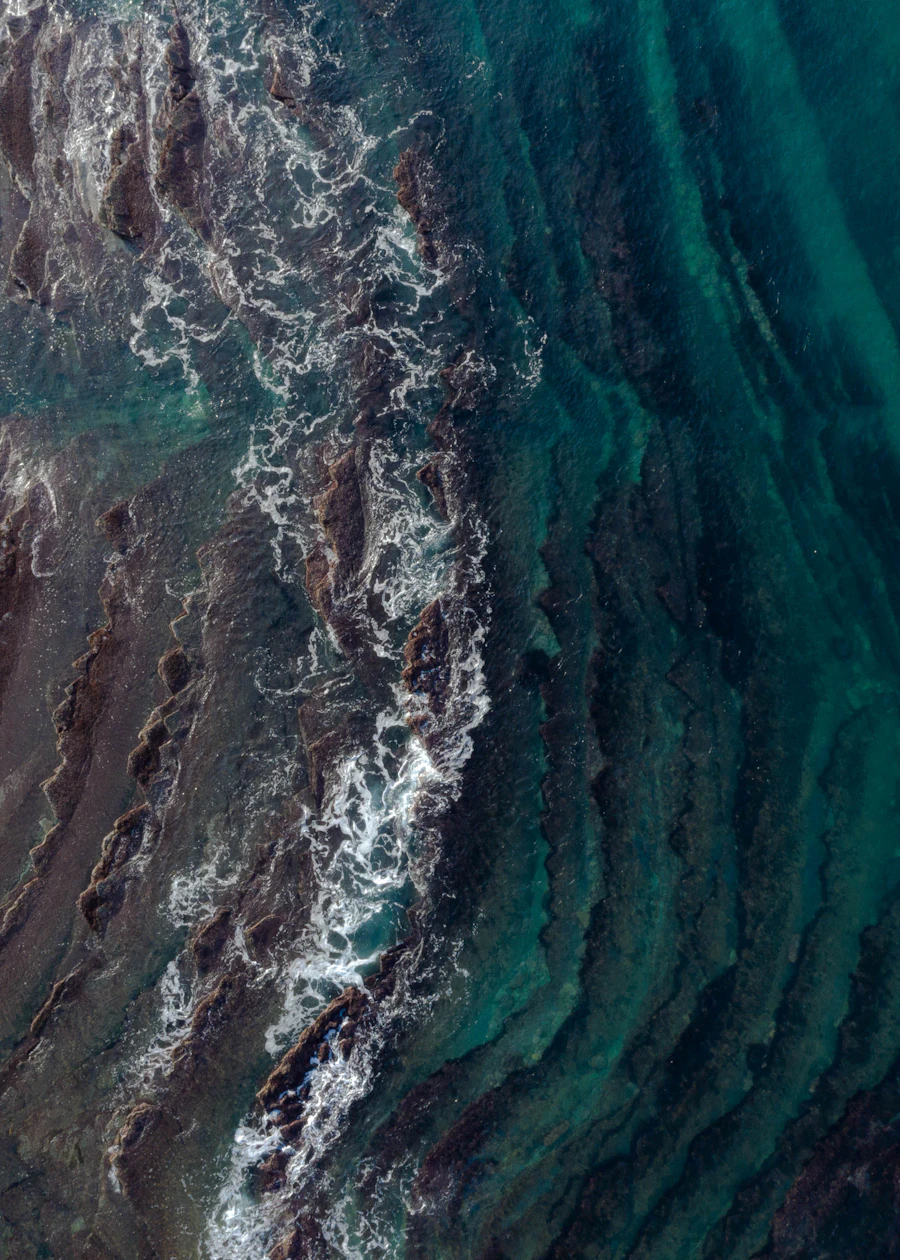 Vibrant aerial view of turquoise sea with receding white foam along rocky lines, revealing layered underwater textures and color gradients.