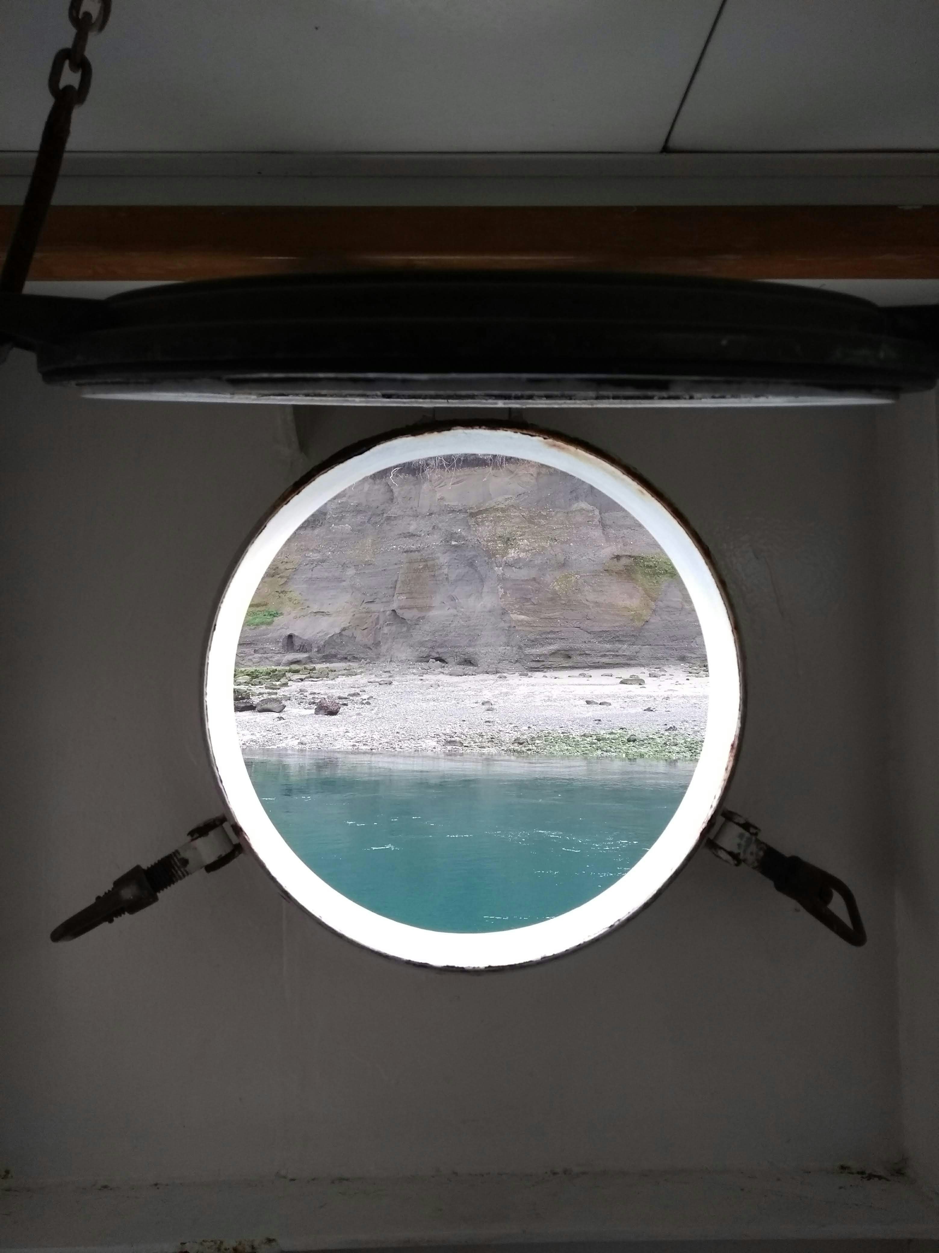 Circular porthole frames turquoise water and a rocky shoreline beyond a ship's interior, capturing a calm maritime seascape.