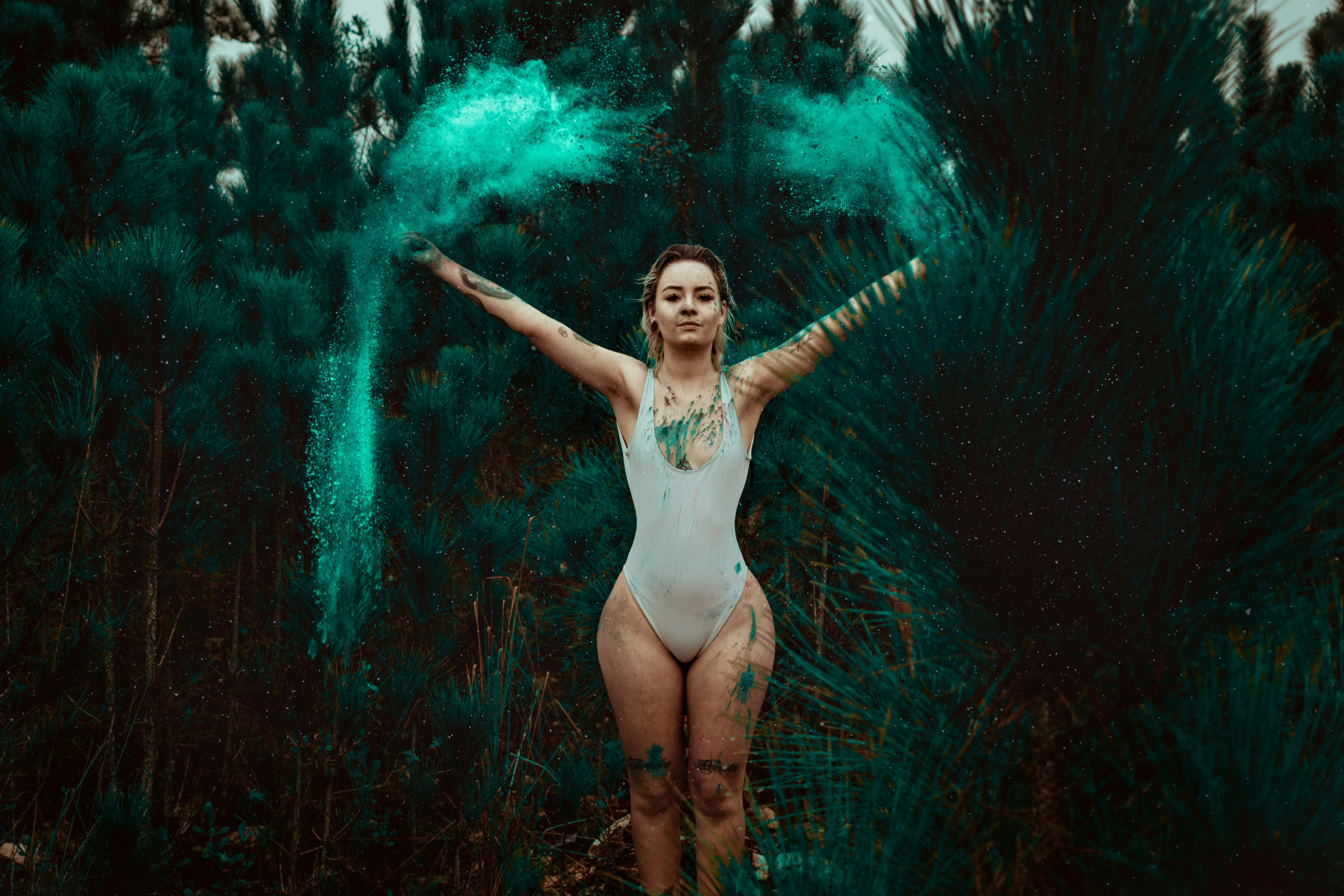 Person in a white swimsuit throws vibrant green powder into the air amidst lush pine trees.