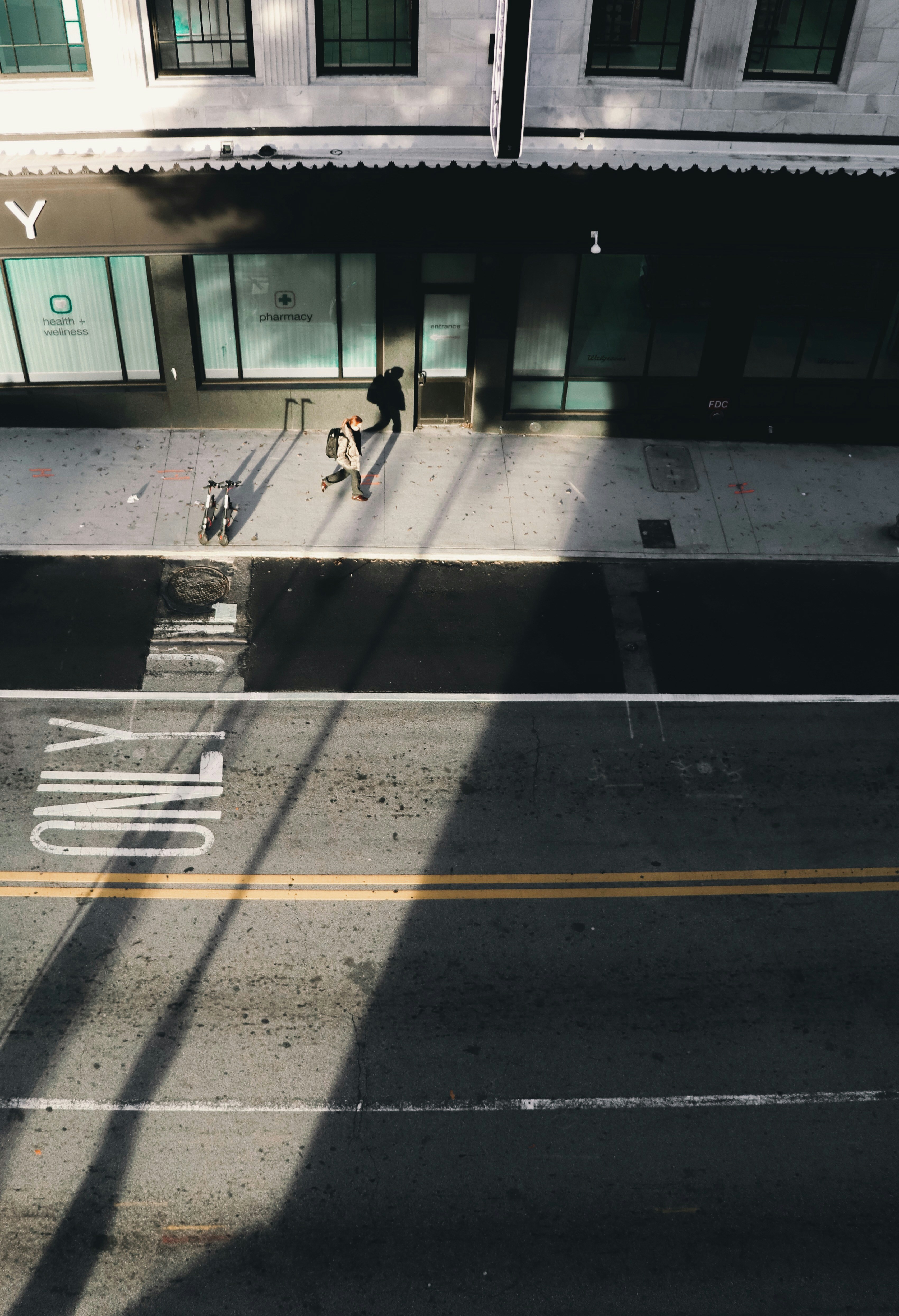 A pedestrian walks alongside a bike on a sunlit urban sidewalk, casting long shadows across the street. The scene captures the interplay of light and architecture.