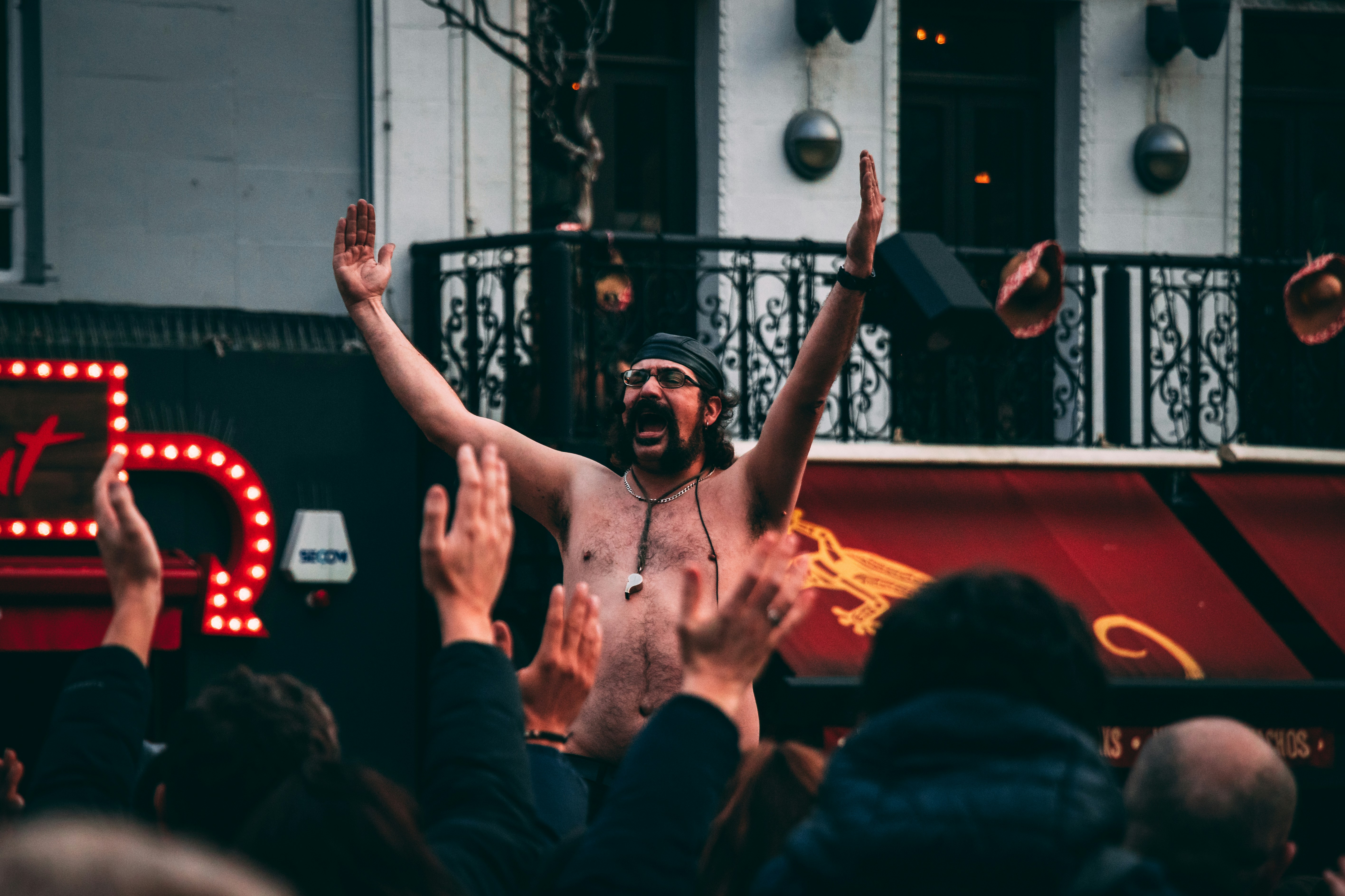 A lively street performer engages an enthusiastic audience, raising his arms in celebration. The backdrop features vibrant signage, adding to the energetic atmosphere.