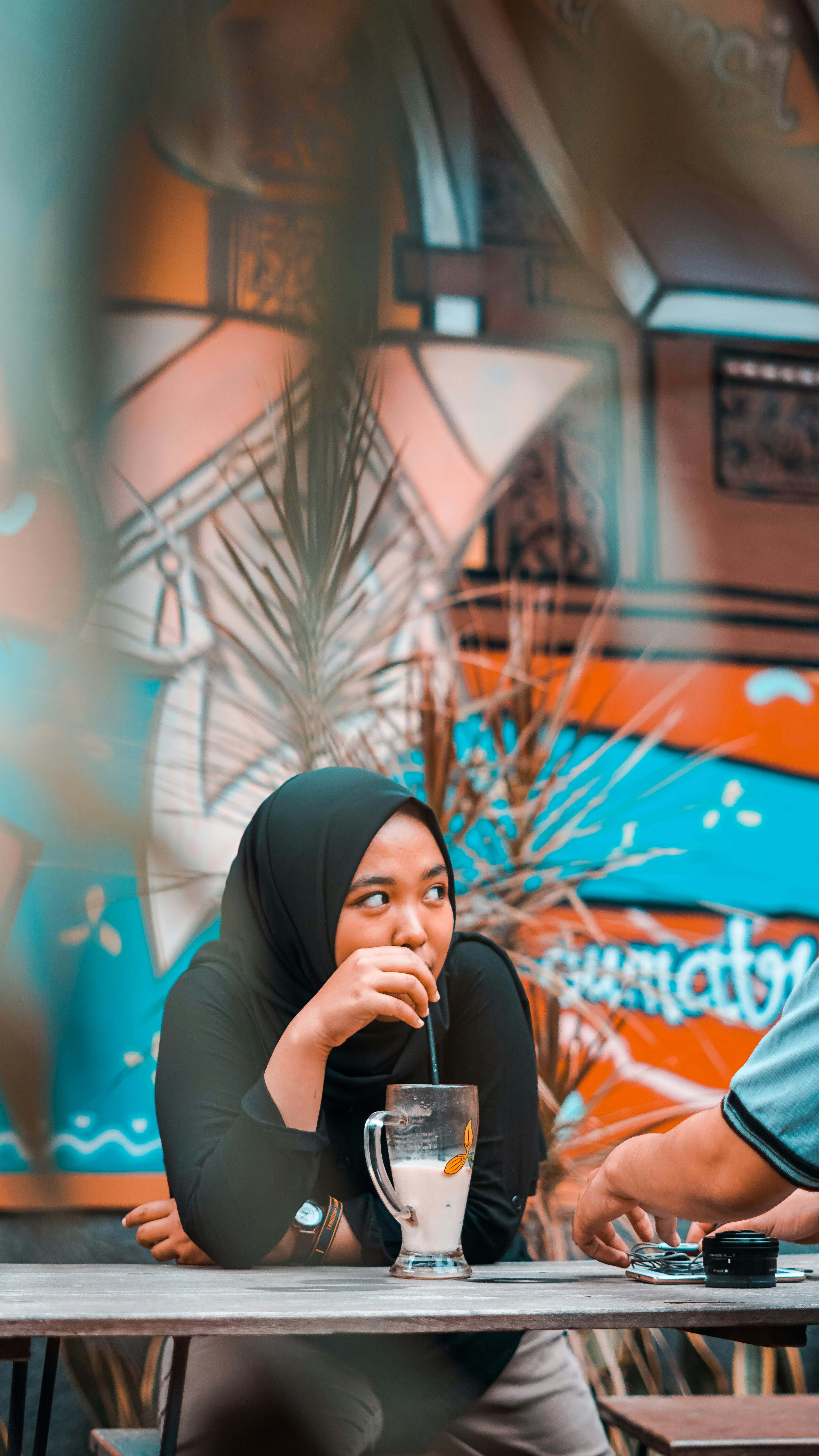 A woman in a black hijab enjoys a drink while seated at a table, surrounded by vibrant mural art and lush greenery.