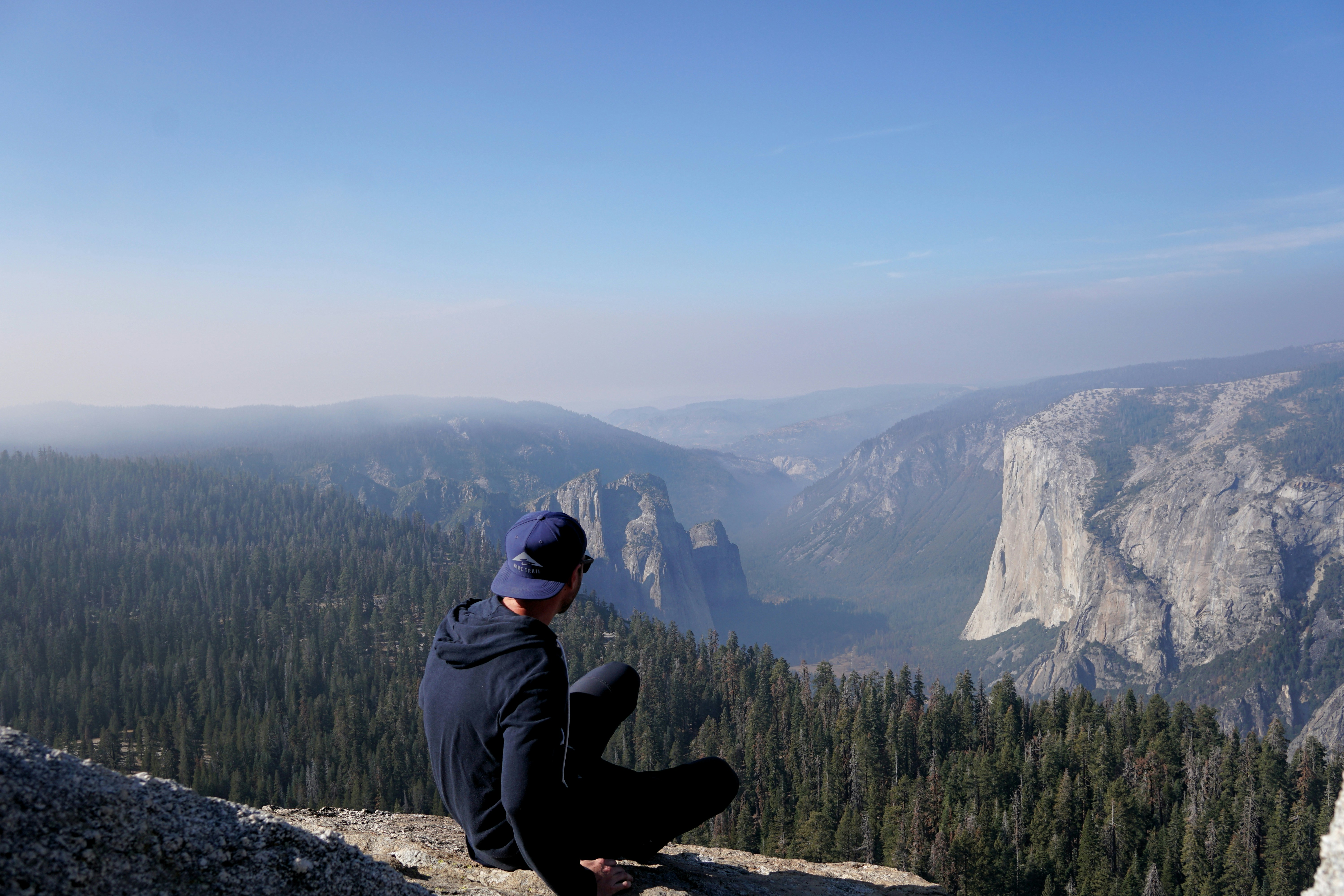 A person sits on a rocky ledge, gazing at the expansive Yosemite Valley framed by towering cliffs and dense forests. The scene captures a moment of introspection in a breathtaking natural landscape.