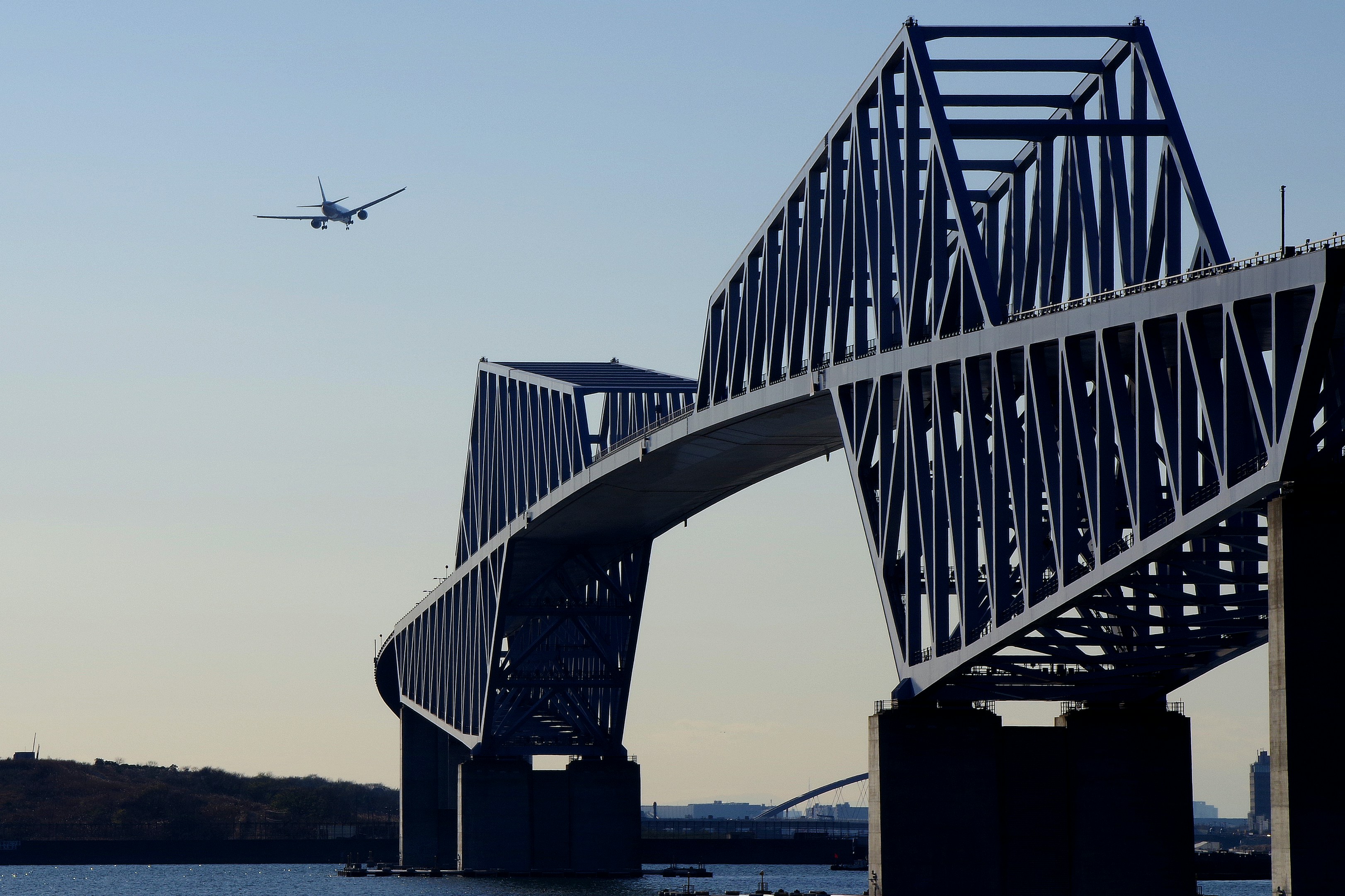 Jetliner approaching over a cobalt-blue steel truss bridge spanning calm water. This photograph highlights the bridge geometry against a clear sky.