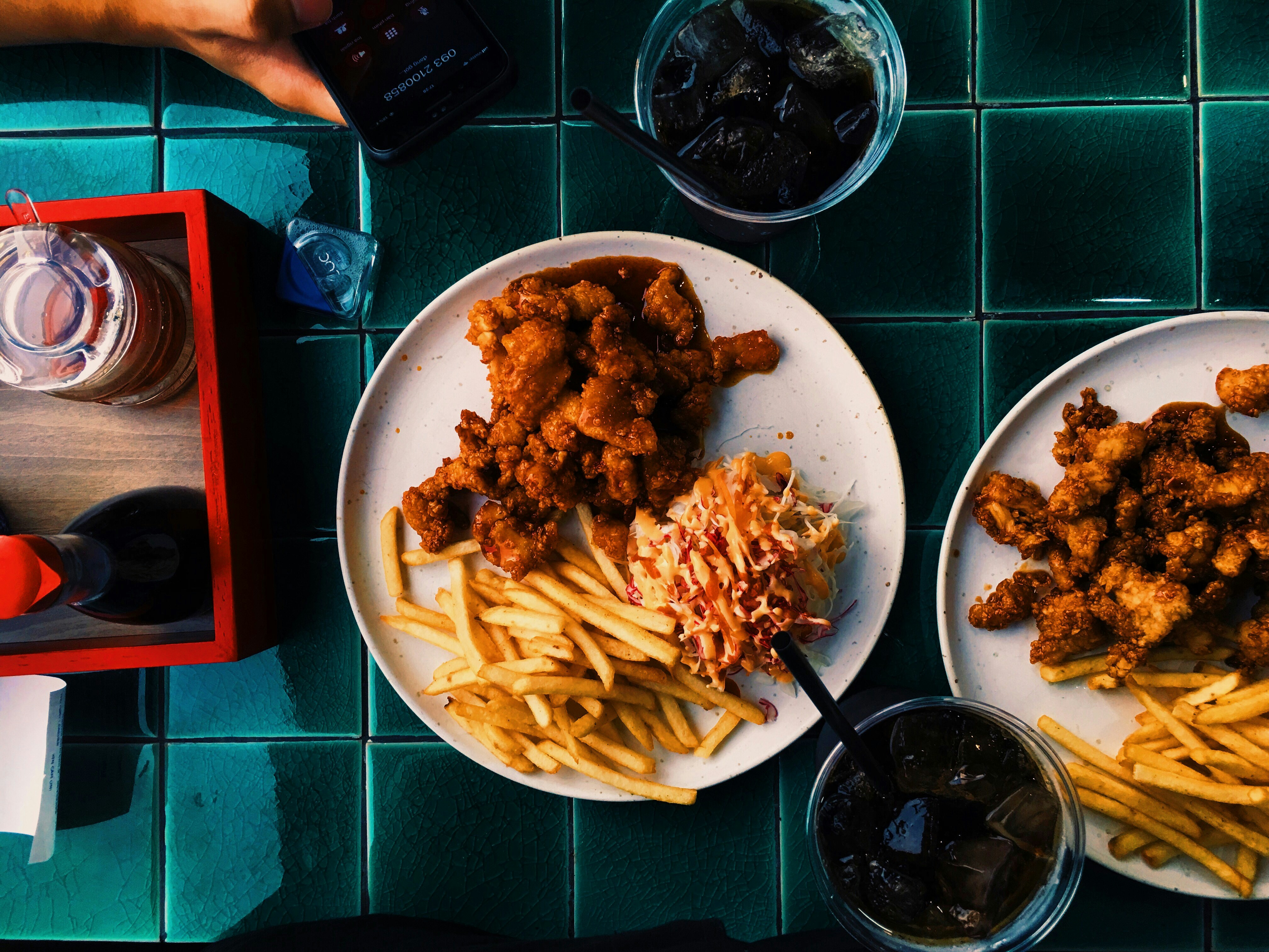 Two plates of crispy fried chicken and fries, accompanied by refreshing drinks, set against a vibrant green tiled table.