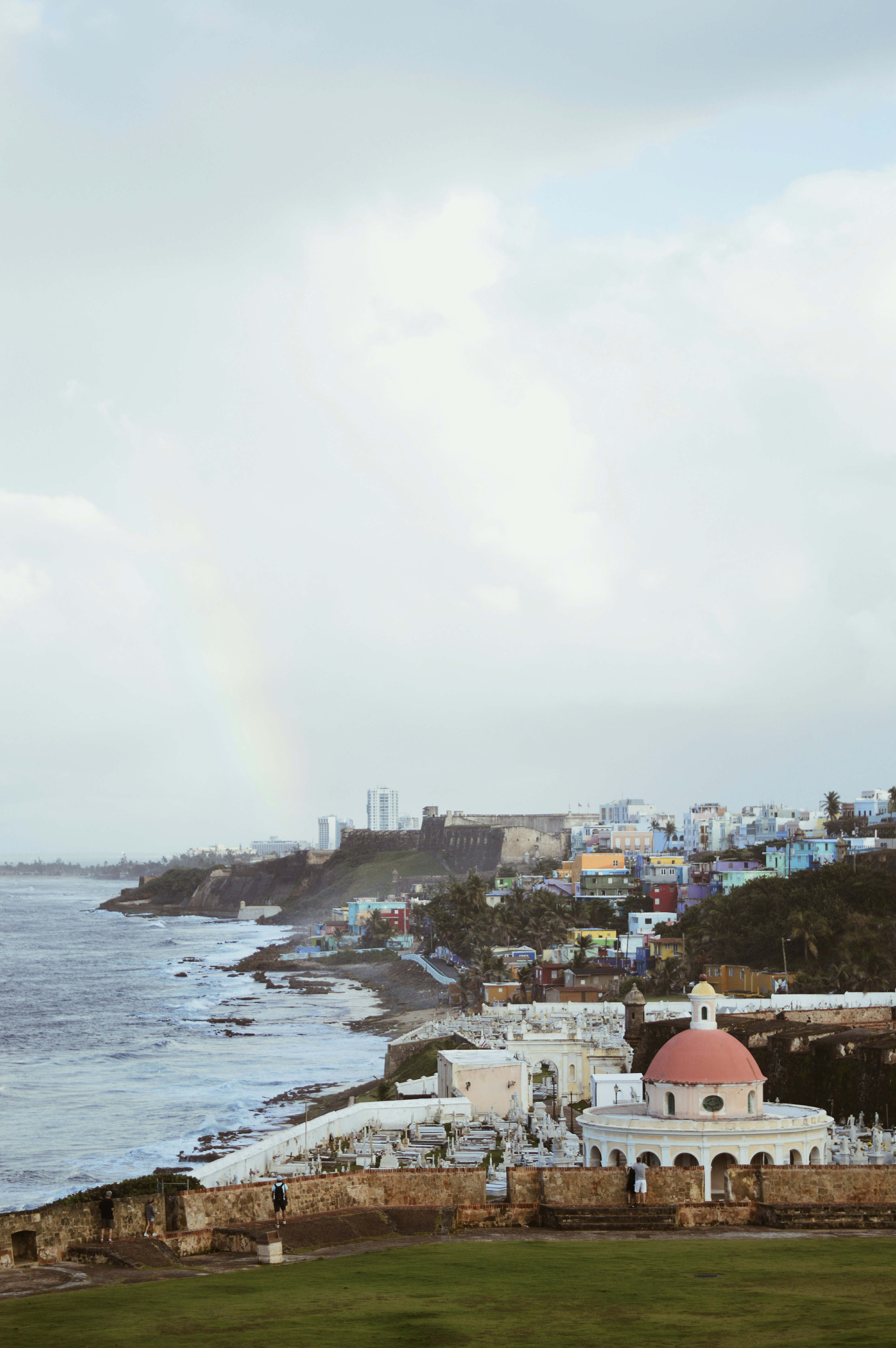 Vibrant coastal landscape featuring colorful buildings and a historic structure with a red dome, set against a dramatic sky and ocean waves. A rainbow adds a touch of magic to the scene.