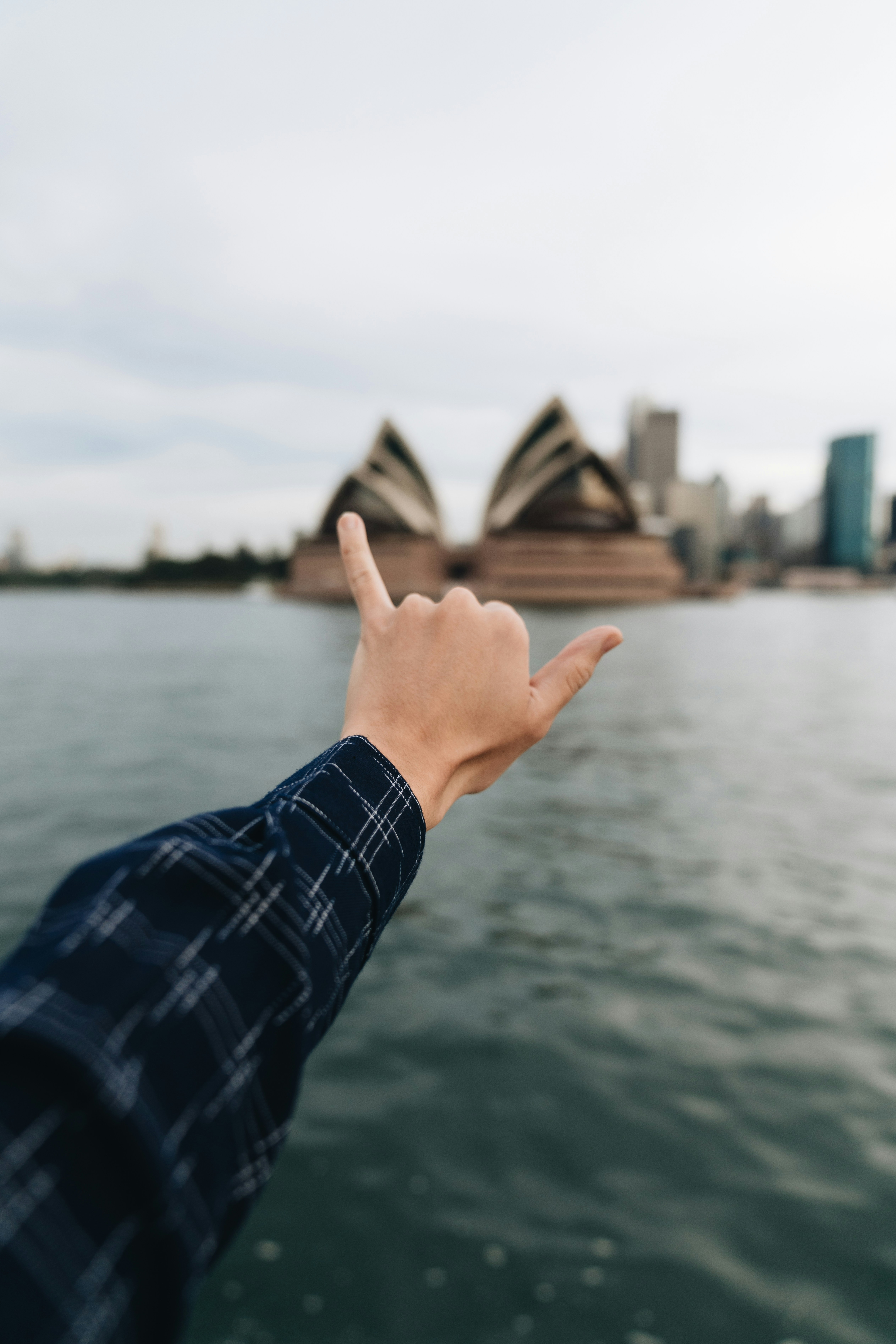 Hand making a shaka sign in front of the Sydney Opera House, with a city skyline in the background.