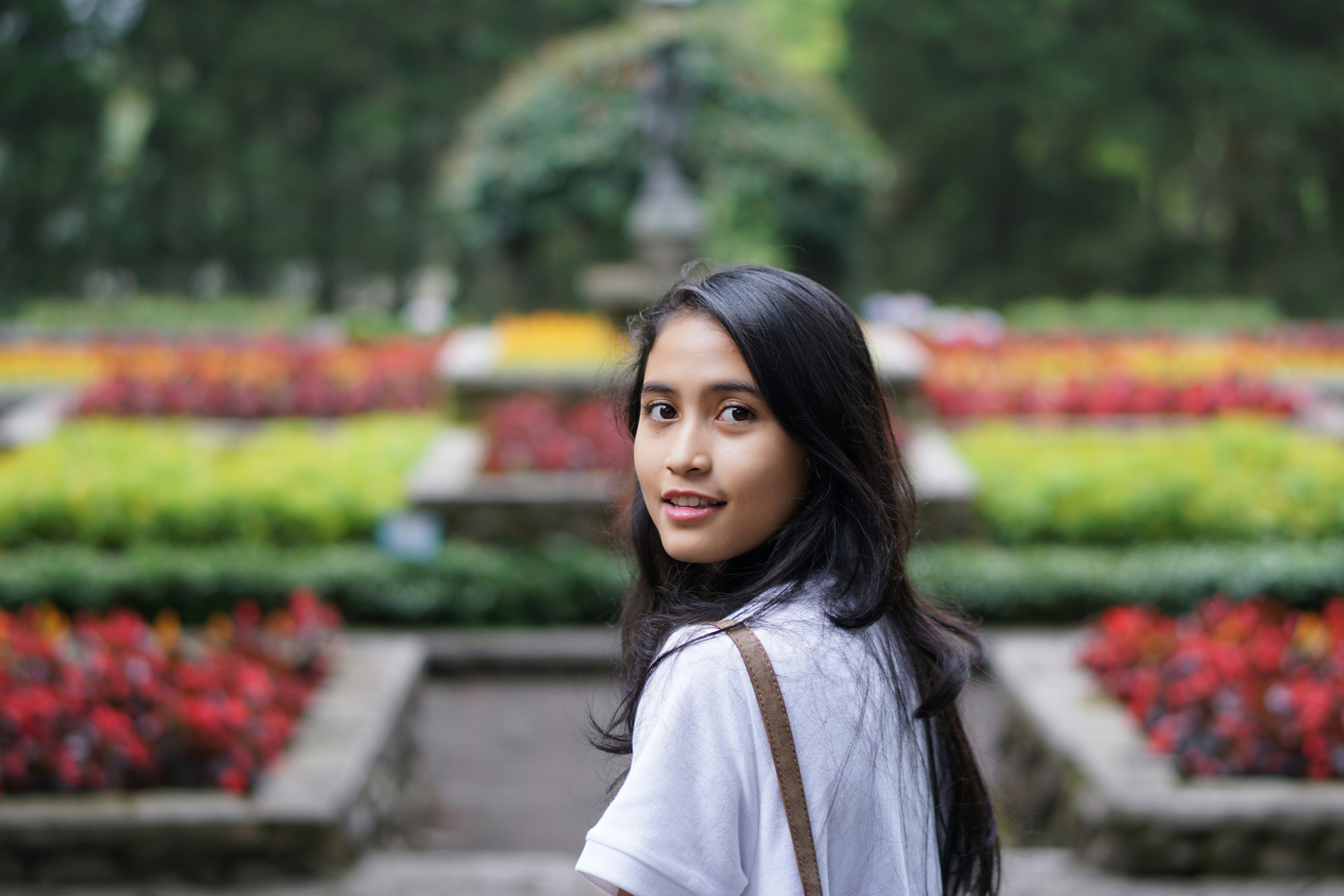A young woman glances back while strolling through a vibrant floral garden, surrounded by a spectrum of blooming flowers.