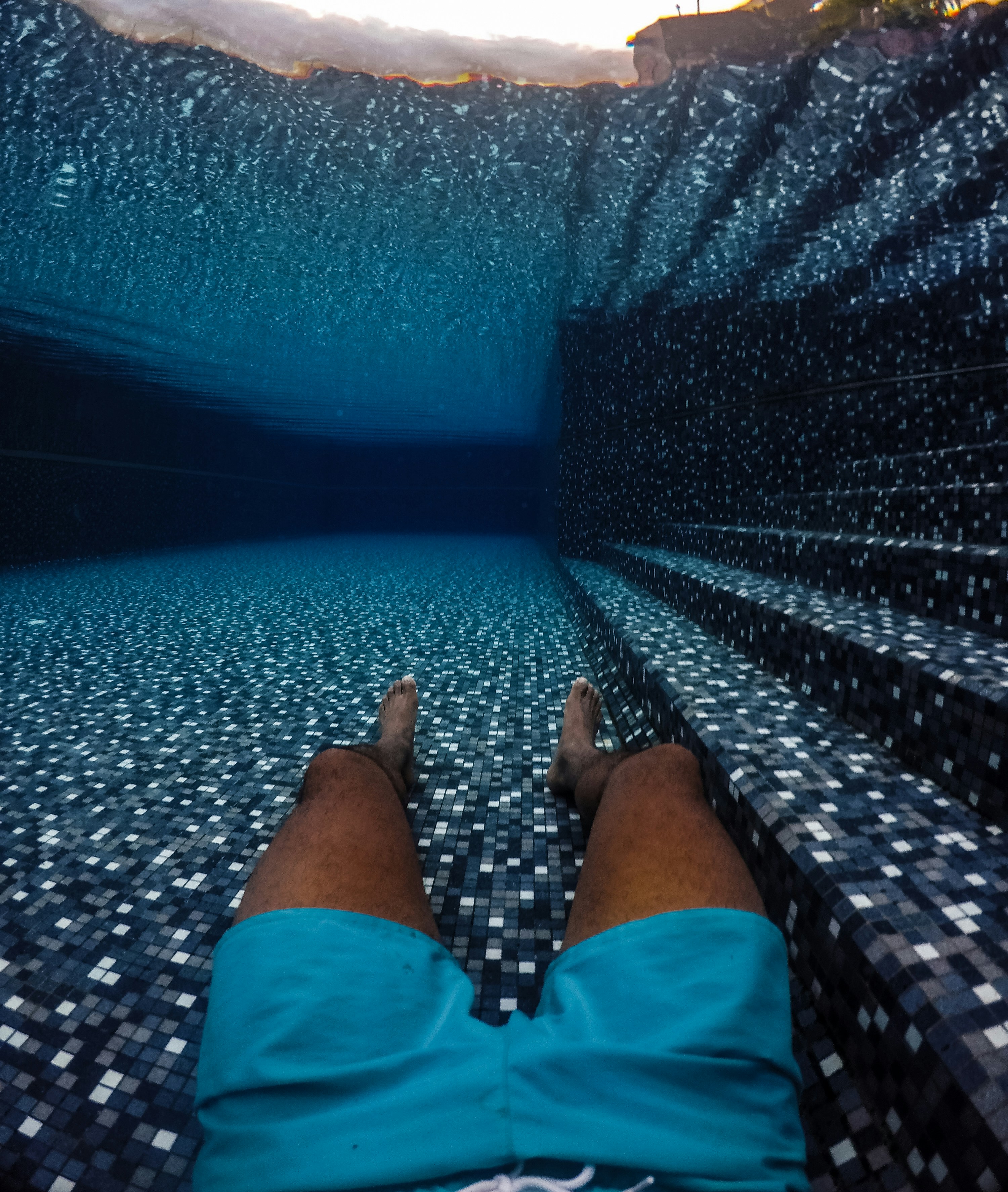 Underwater photograph of a swimmer lounging with legs extended toward the far wall, captured from a submerged vantage in a blue-tiled pool.