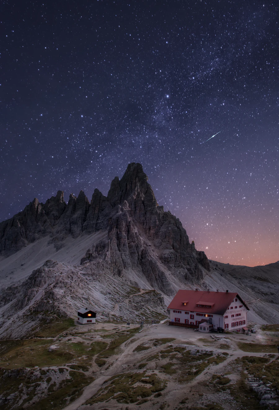 A rugged mountain peak rises above an alpine hut with a red roof under a dense star-filled night sky. A faint meteor streaks across the upper right as dusk glows on the horizon.