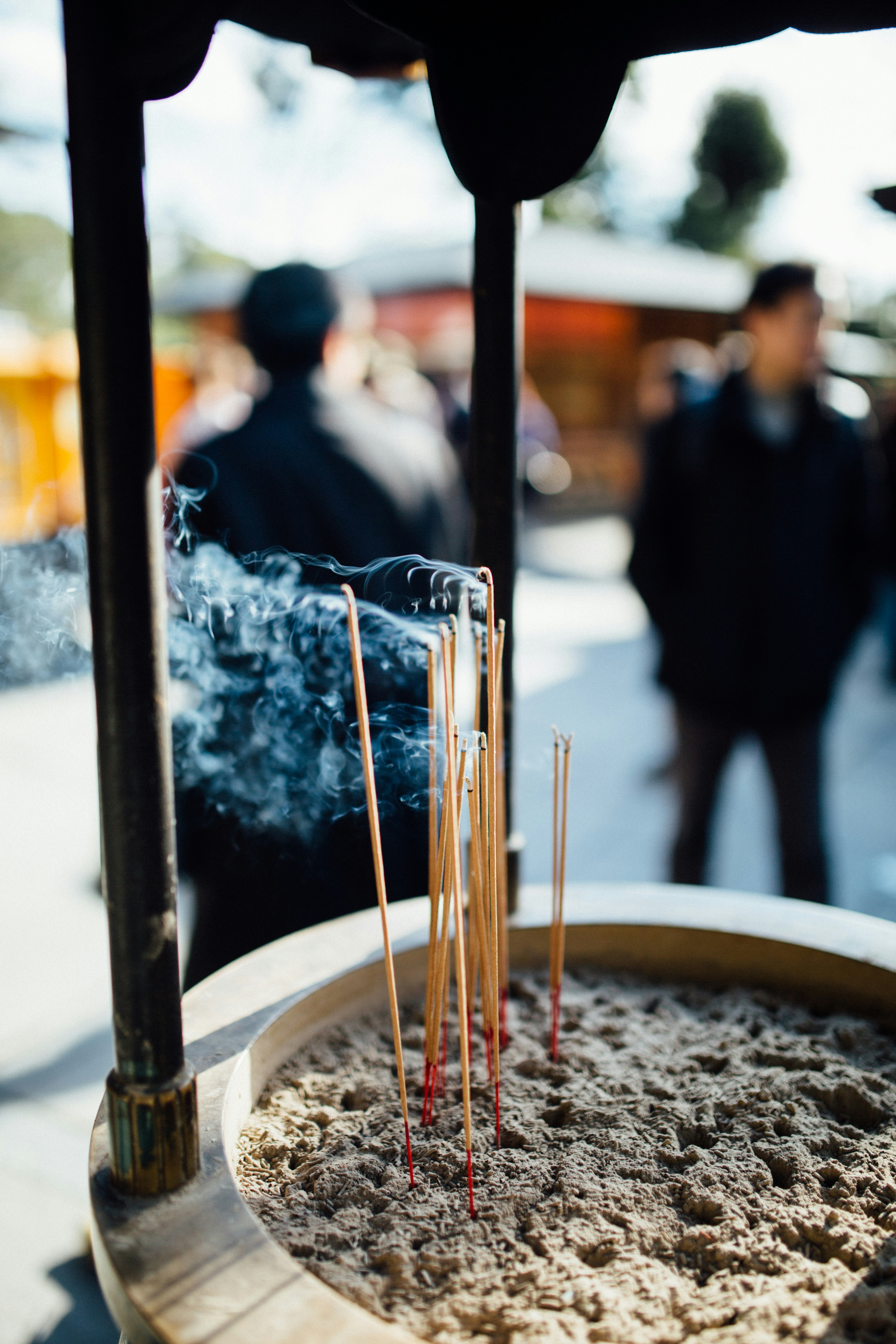 Incense sticks standing in sandy receptacle, with smoke curling upwards in a serene outdoor setting.