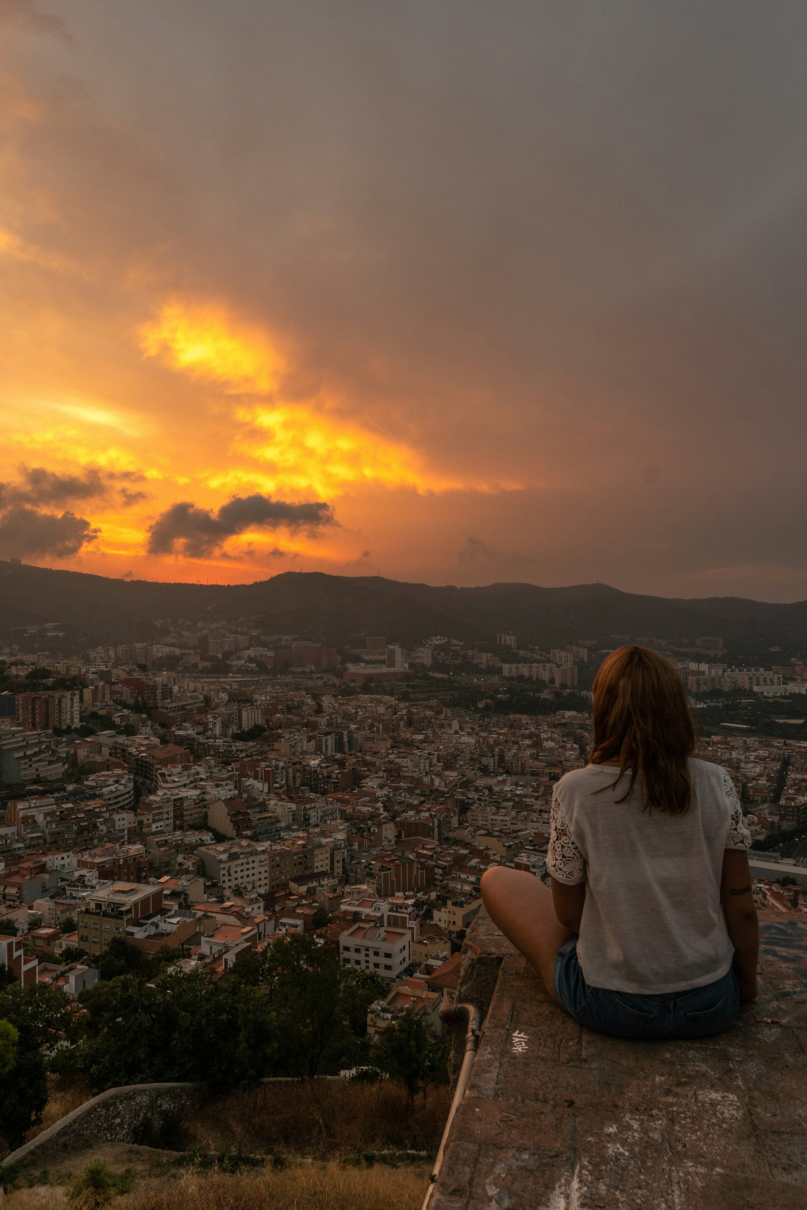 A woman gazes over a sprawling city as a vibrant sunset casts warm hues across the sky. The scene captures a moment of reflection and tranquility.