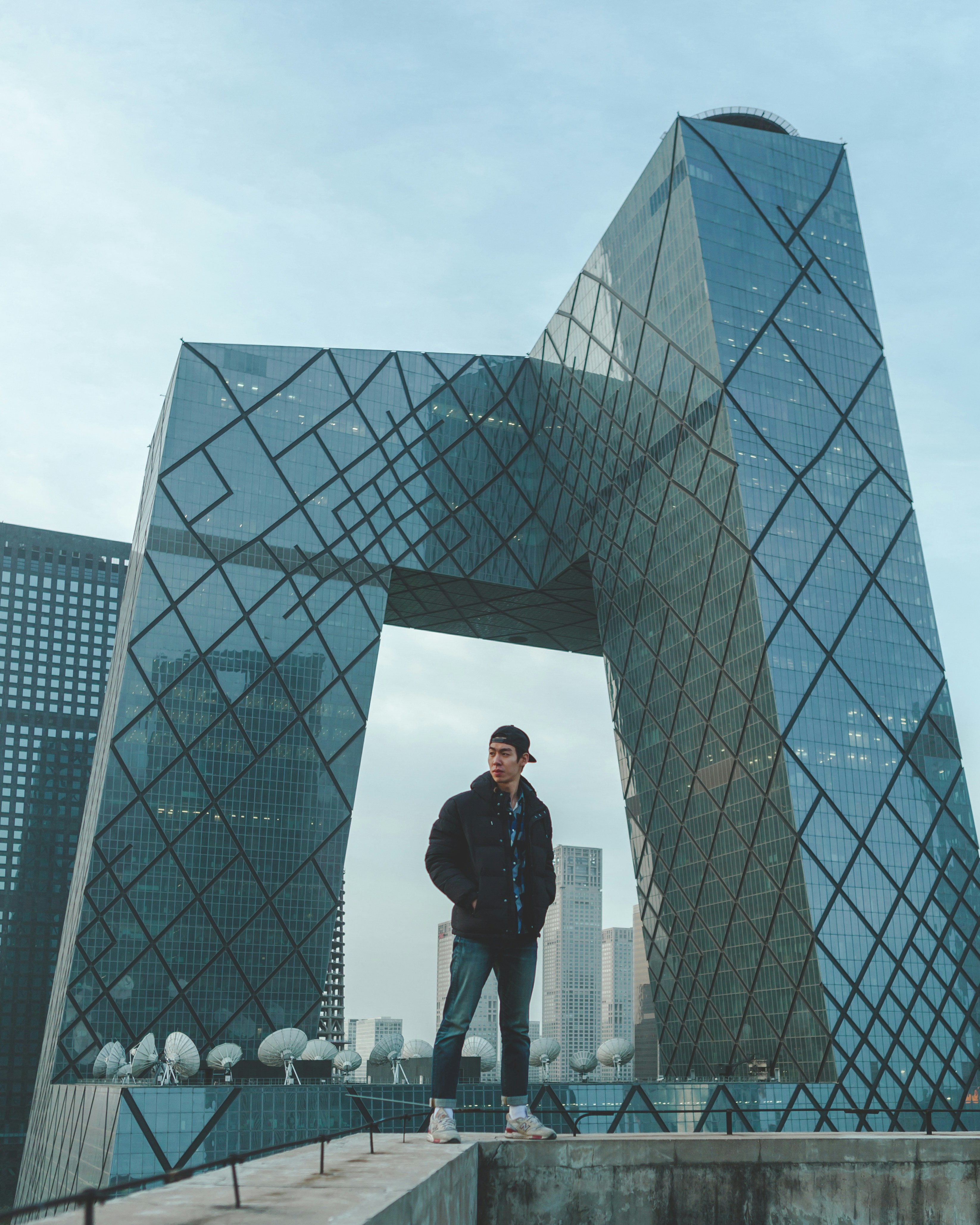 A figure stands confidently atop a ledge, framed by the striking architecture of the CCTV Headquarters in Beijing. The geometric design reflects the city’s innovative spirit.