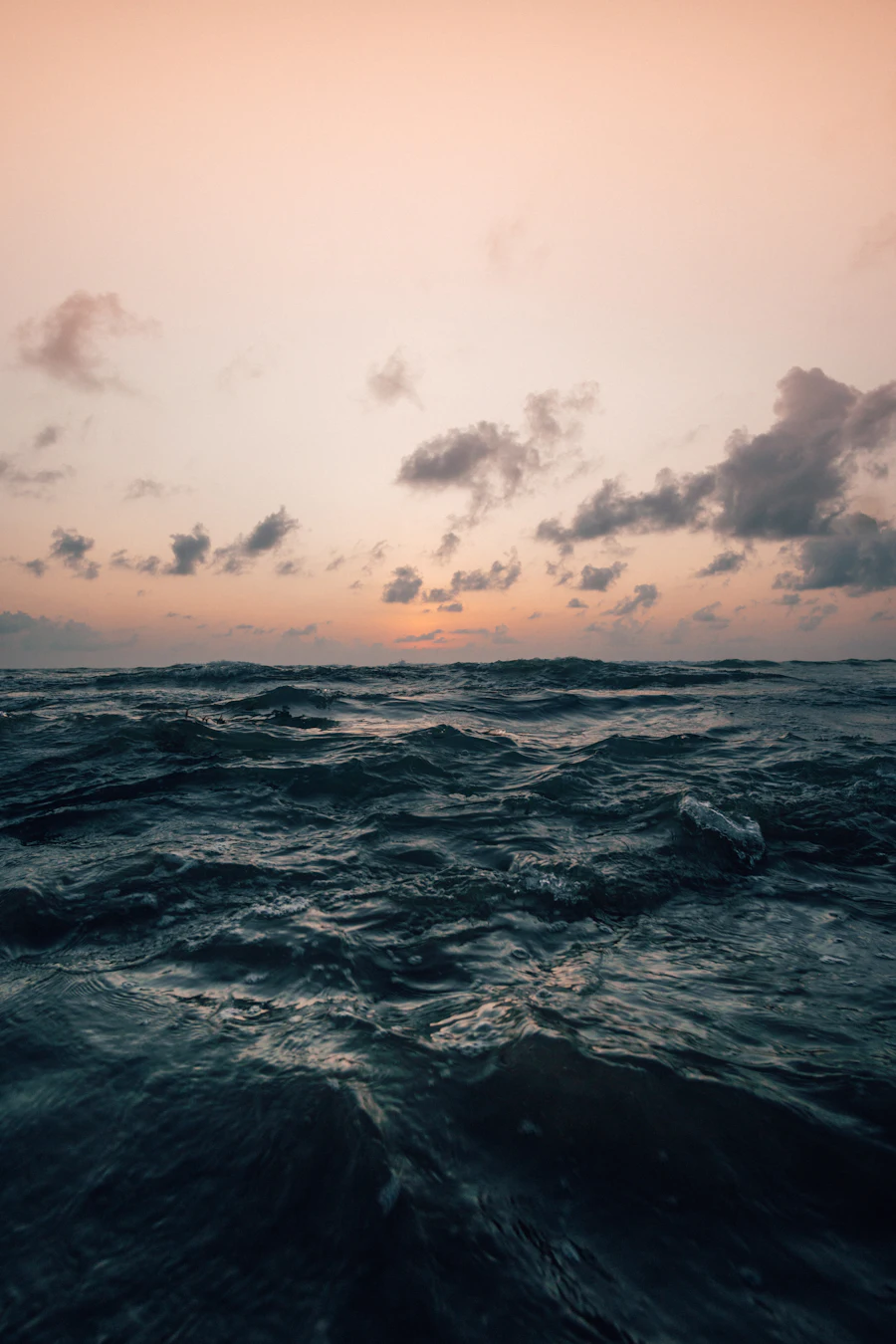 Choppy ocean waves fill the foreground beneath a pastel sunset sky with scattered clouds. The horizon sits low as warm light fades into deep blue water.