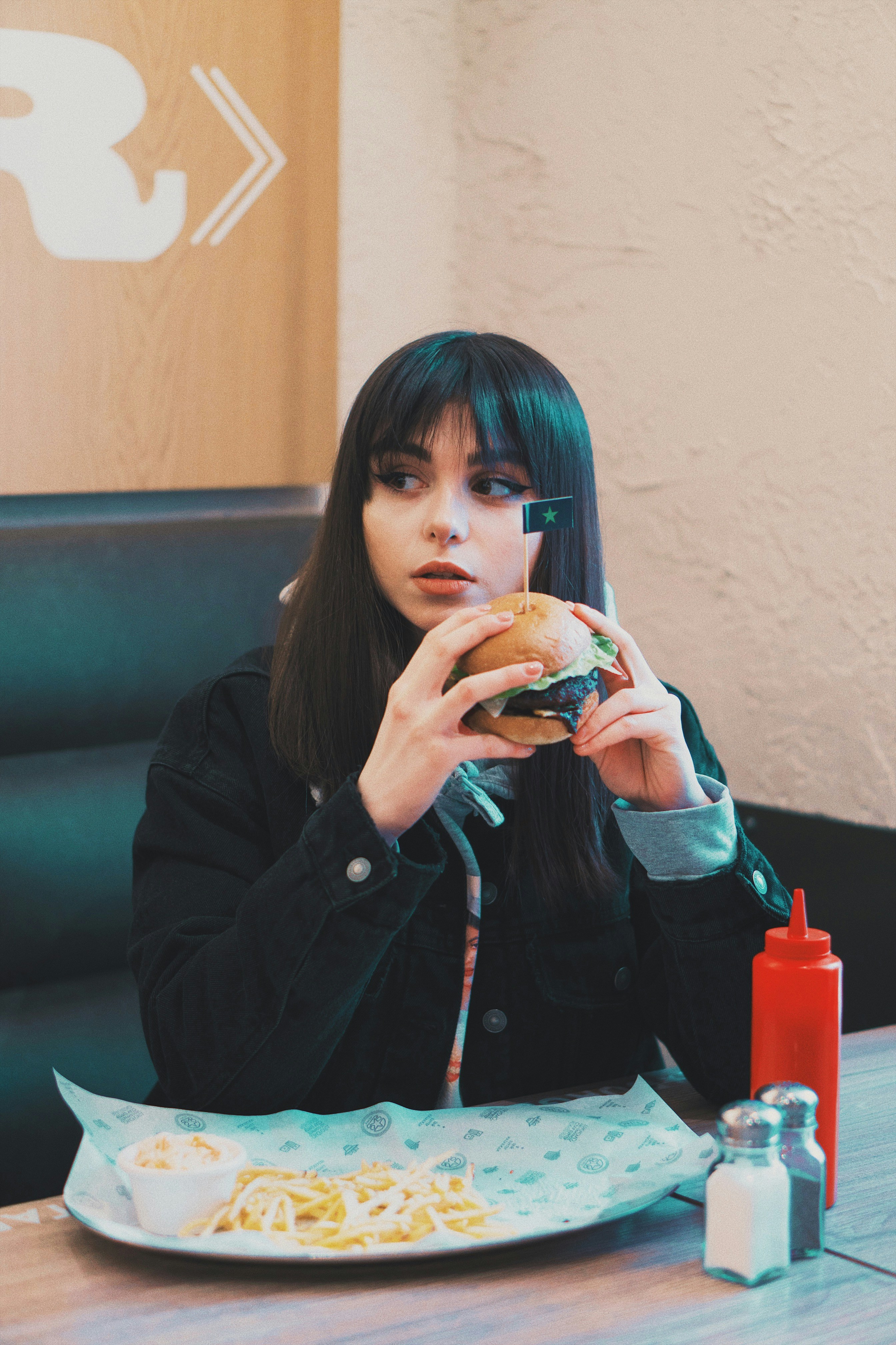 Young woman holding a burger with a flag, seated at a diner table with fries and condiments nearby.