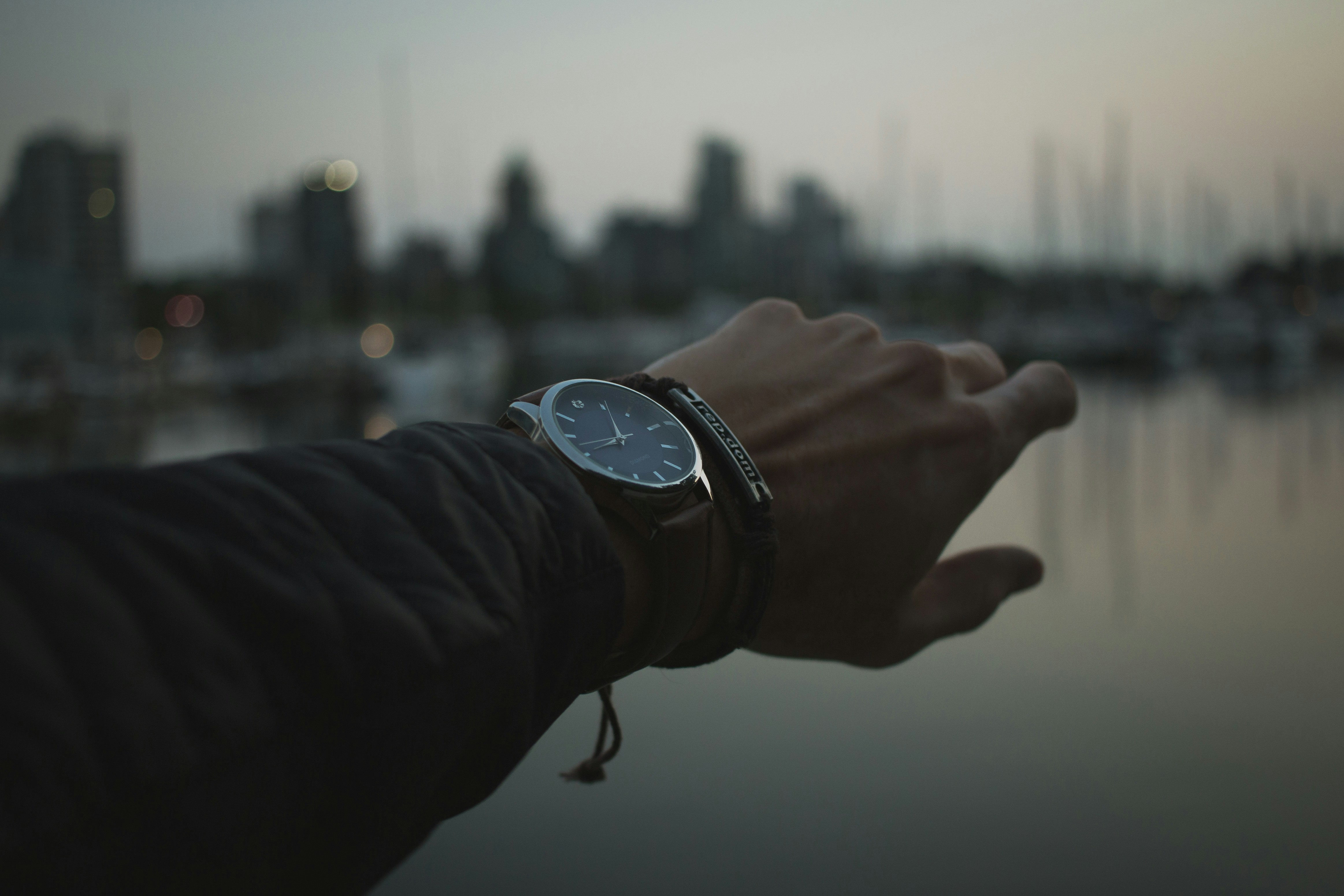 A hand reaching towards the horizon with a stylish watch visible, set against a blurred marina backdrop at dusk.