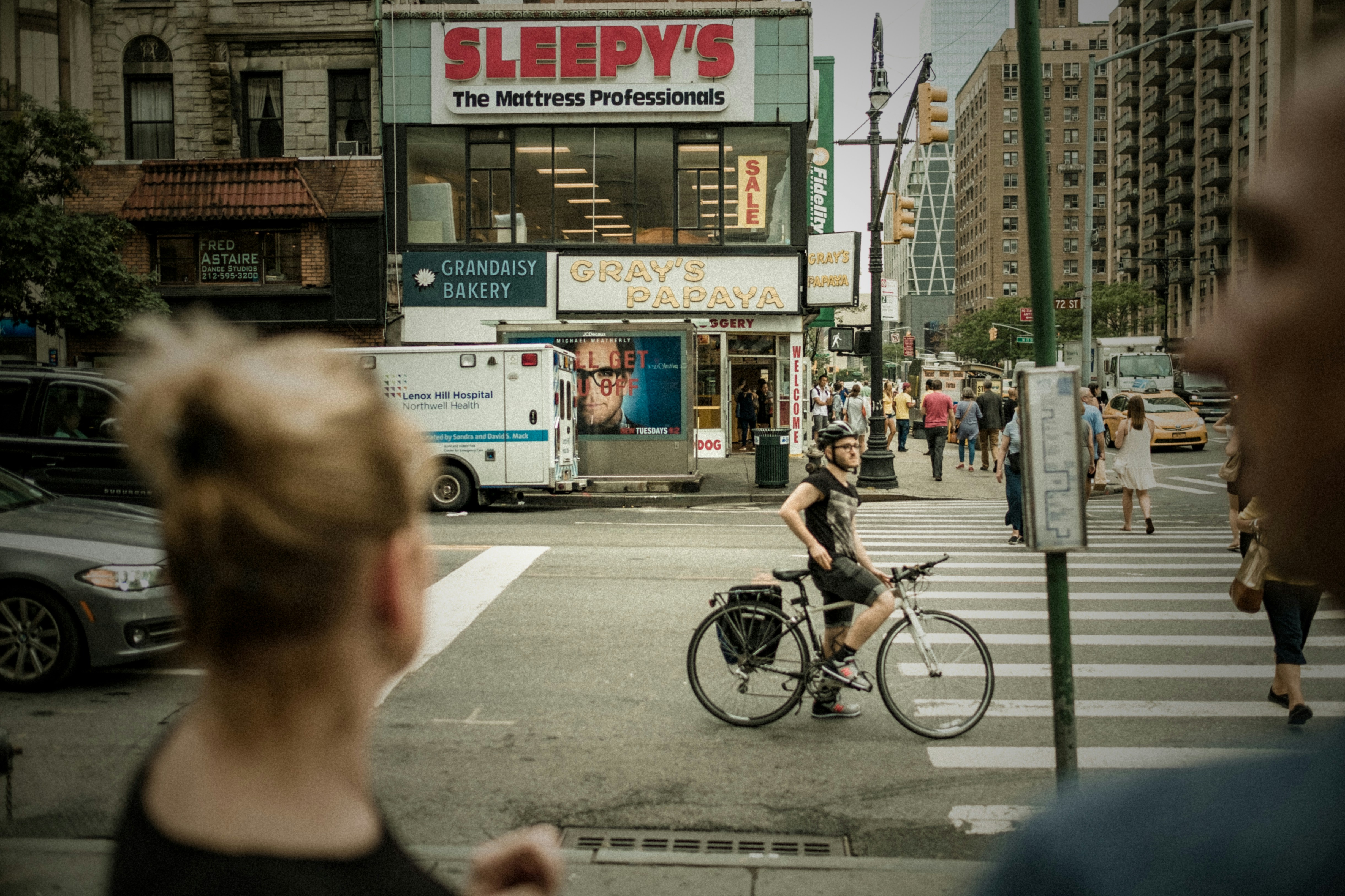 Cyclist pauses at a busy New York City intersection with iconic storefronts in the background.