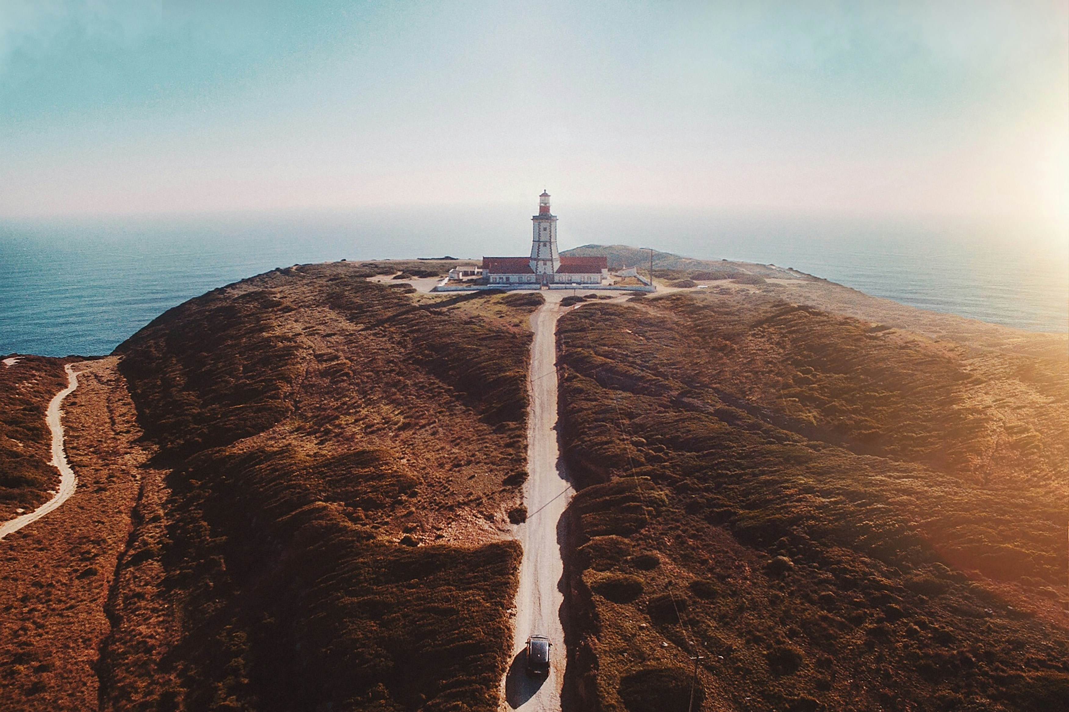 Lighthouse perched on a hilltop with a winding path leading to it, surrounded by ocean views under a clear sky.