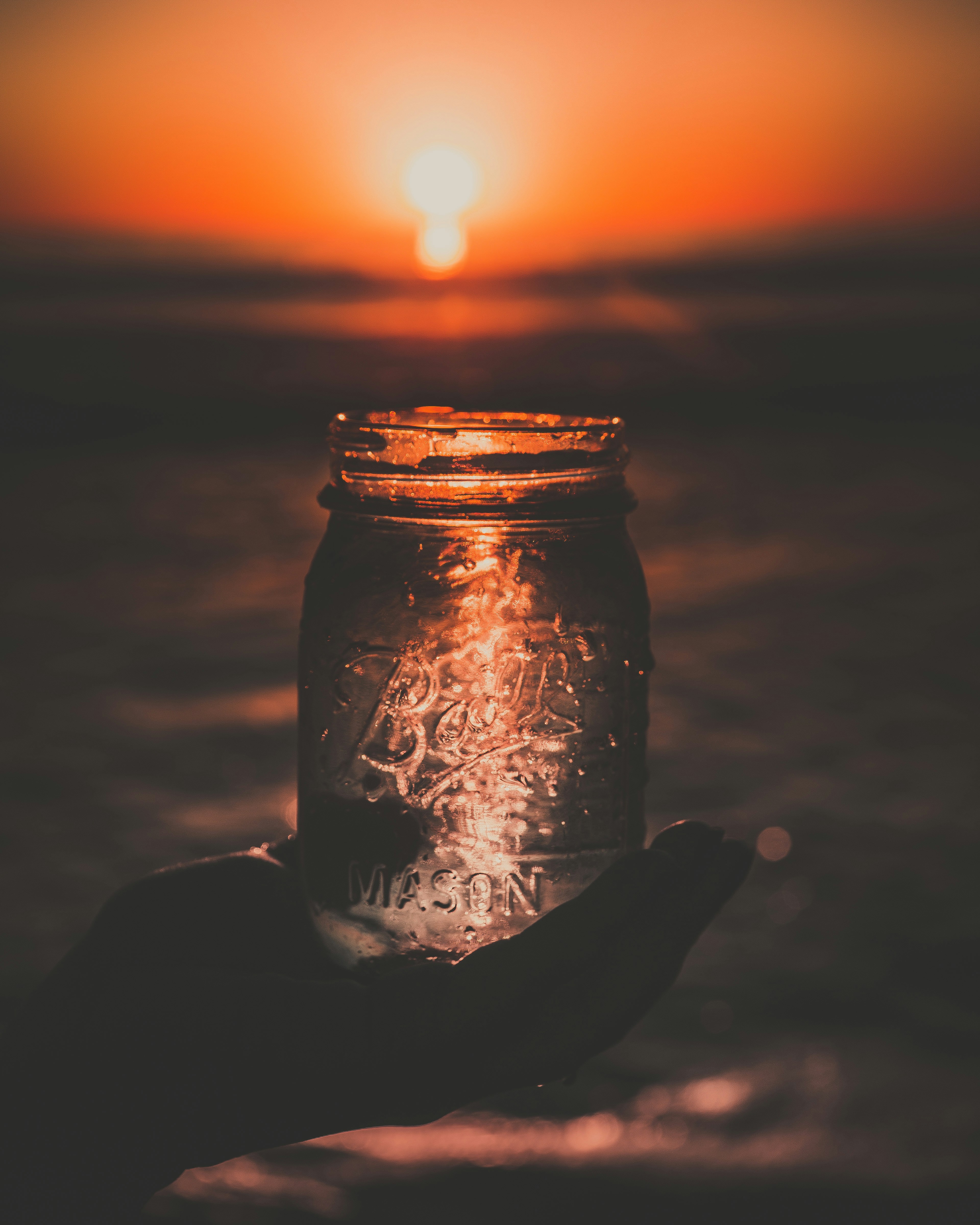 Hand holding a mason jar filled with water, reflecting the vibrant hues of a sunset over the ocean.