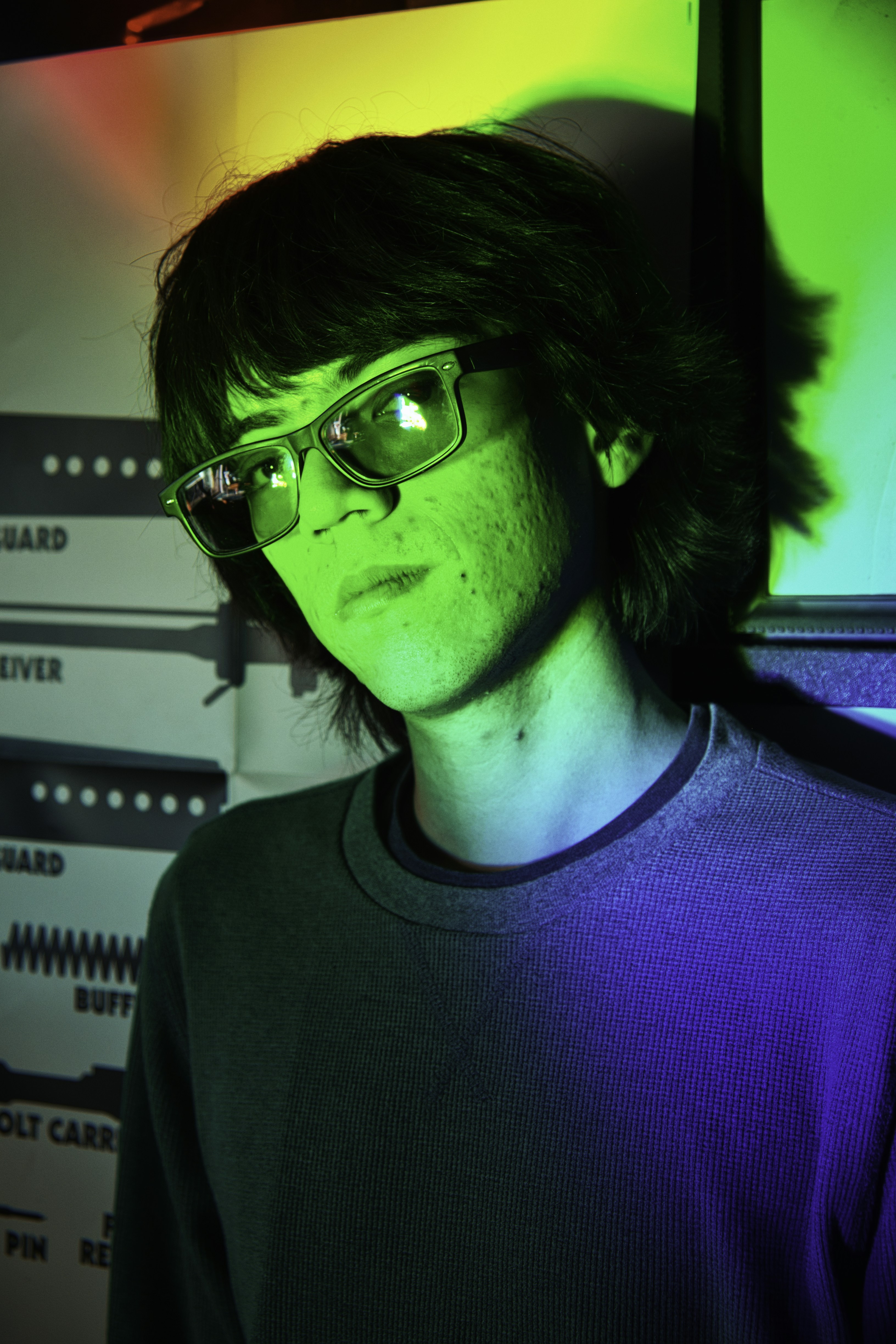 A young man with glasses stands against a backdrop illuminated by vibrant colored lights, creating a striking contrast with his features.