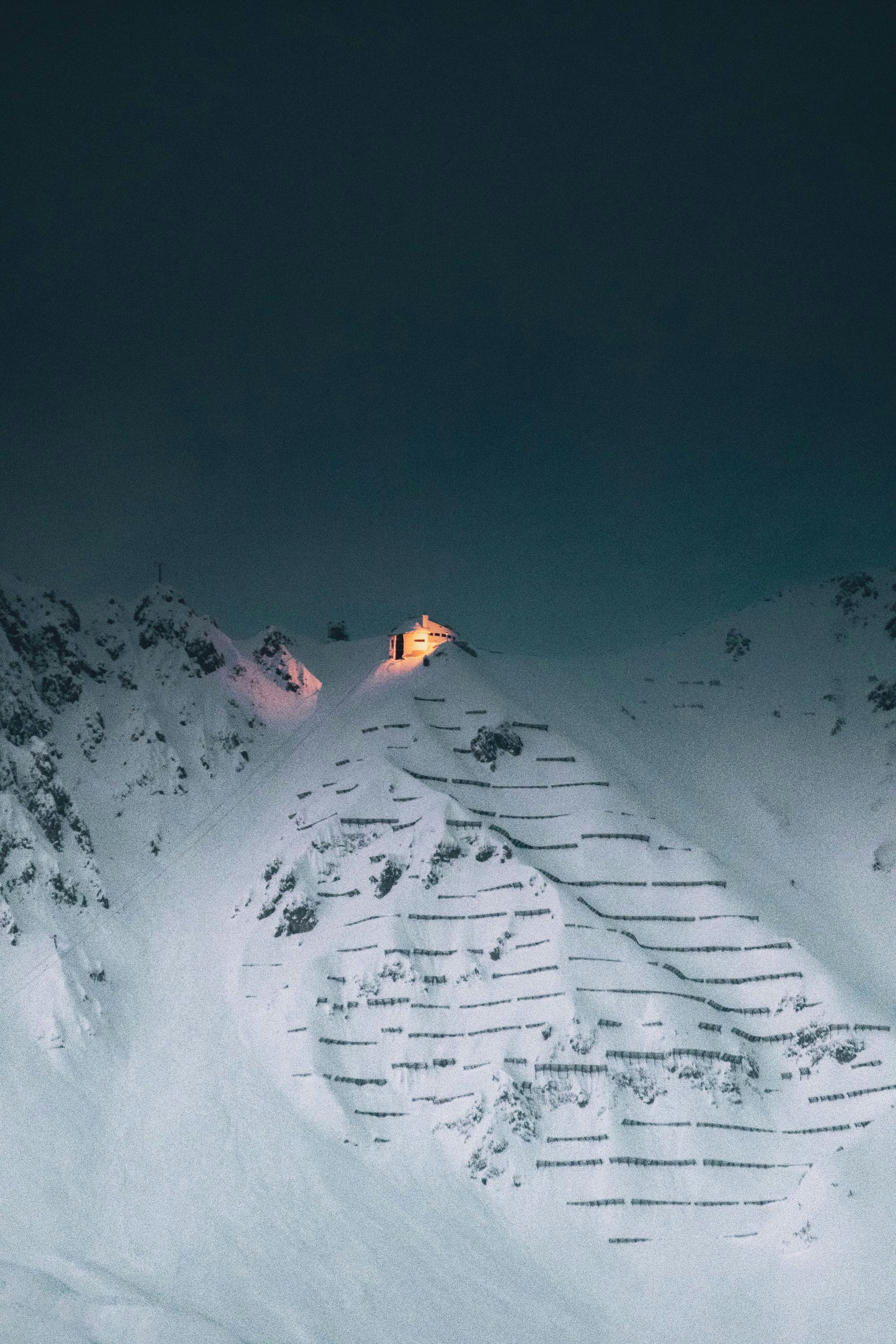 Snow-covered mountain with a distant cabin illuminated at dusk.