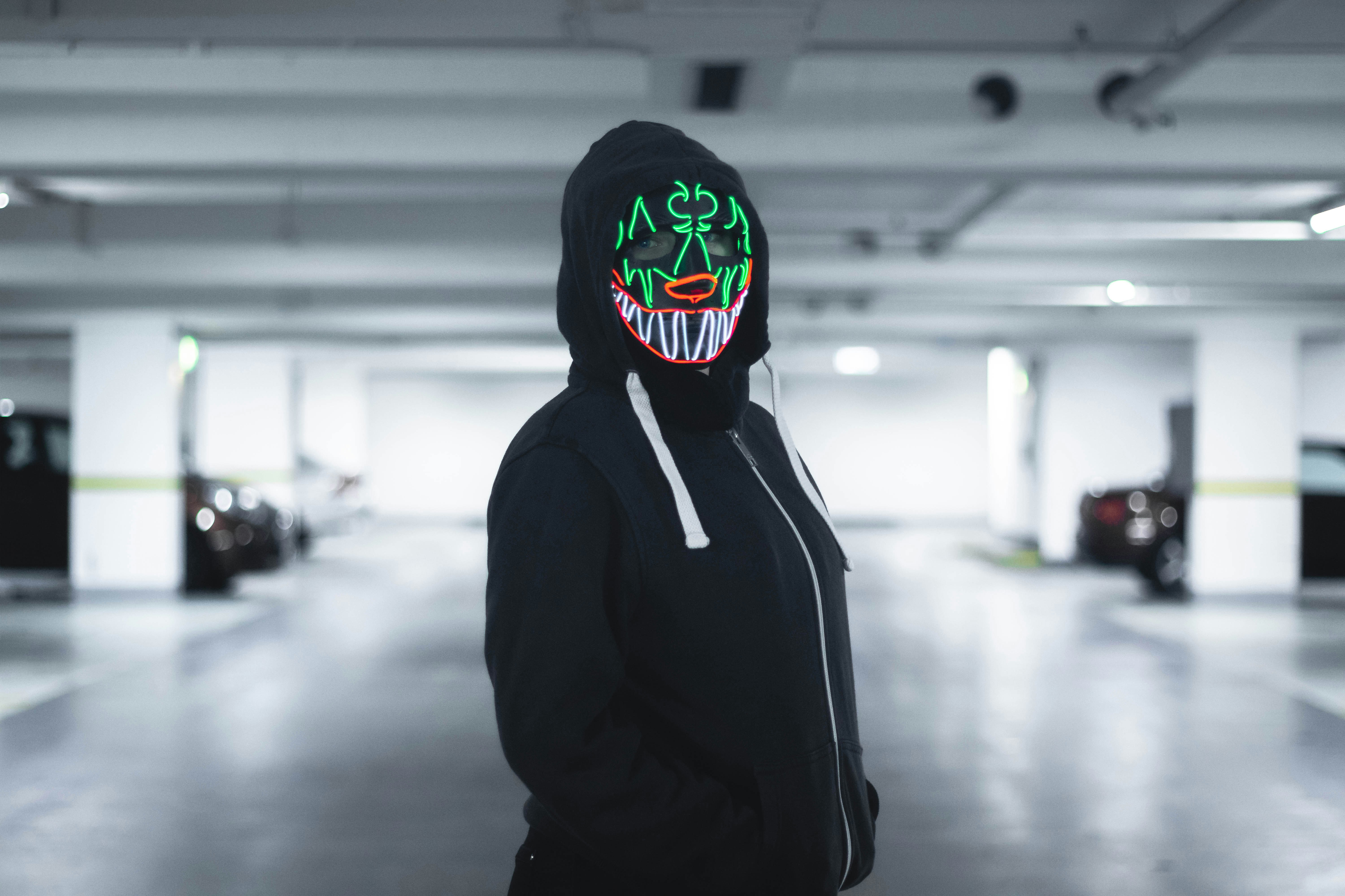 A figure in a dark hoodie stands in a dimly lit parking garage, their face illuminated by a vibrant neon mask design.