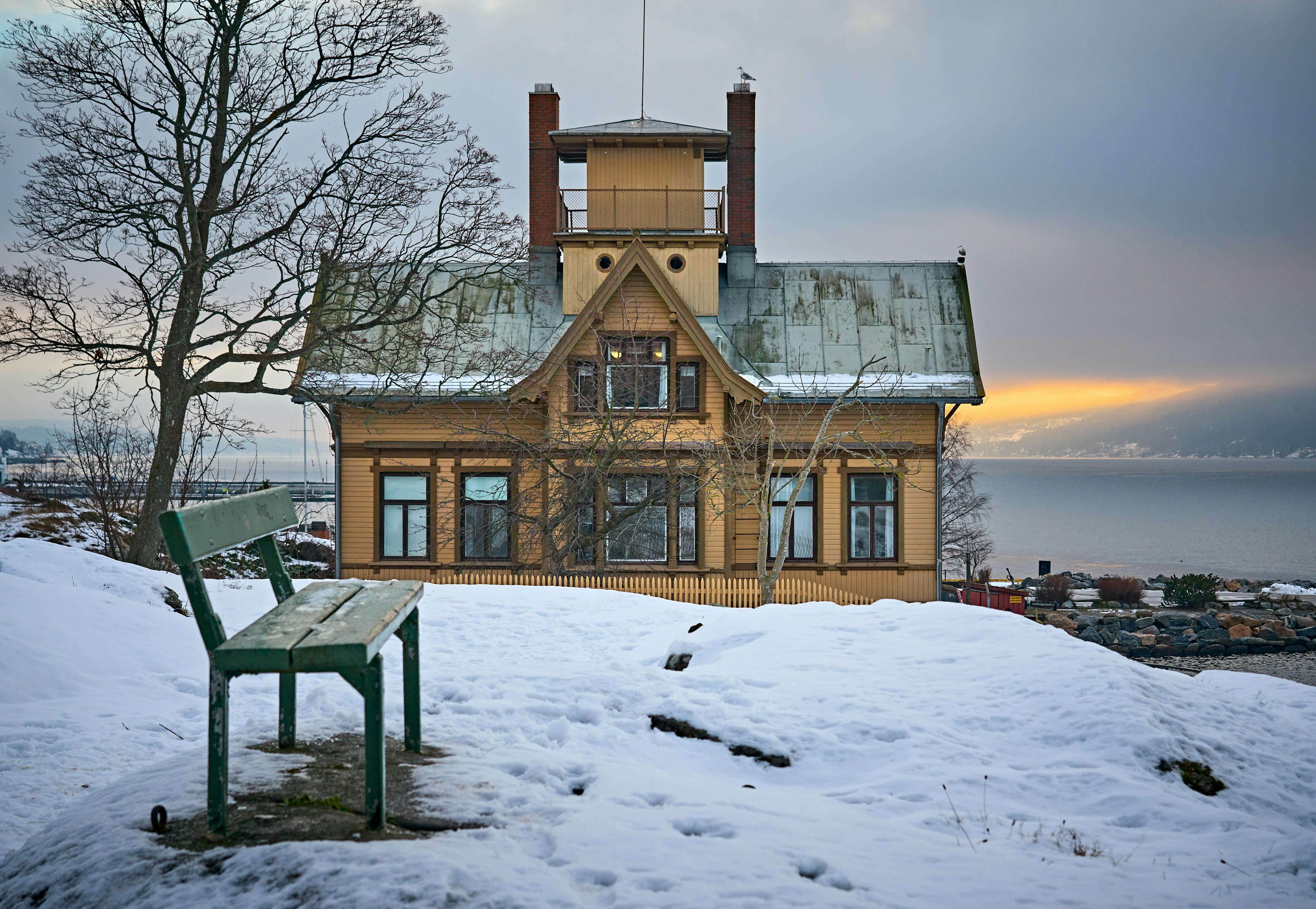 Charming wooden house with a green bench in front, surrounded by snow and a tranquil waterfront at dusk.