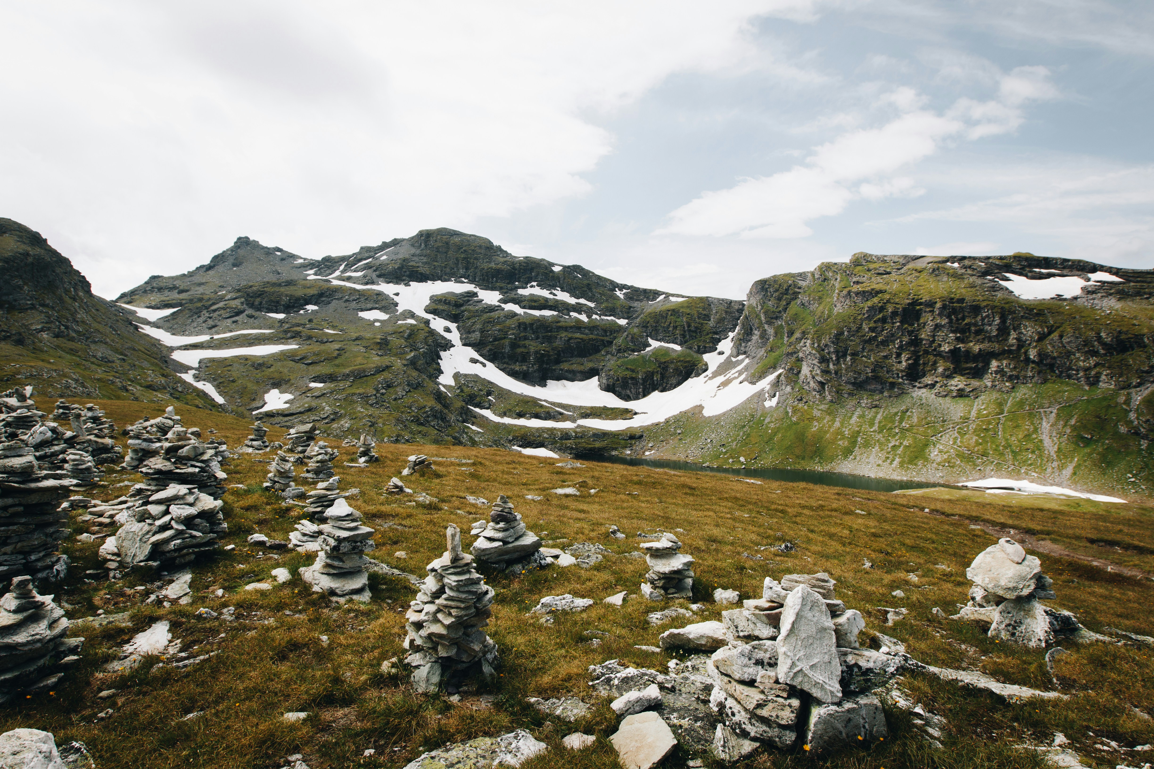 A serene landscape featuring carefully stacked stone cairns amidst a lush green valley and rugged mountains, hinting at human presence in the wild. Snow patches accentuate the natural beauty.