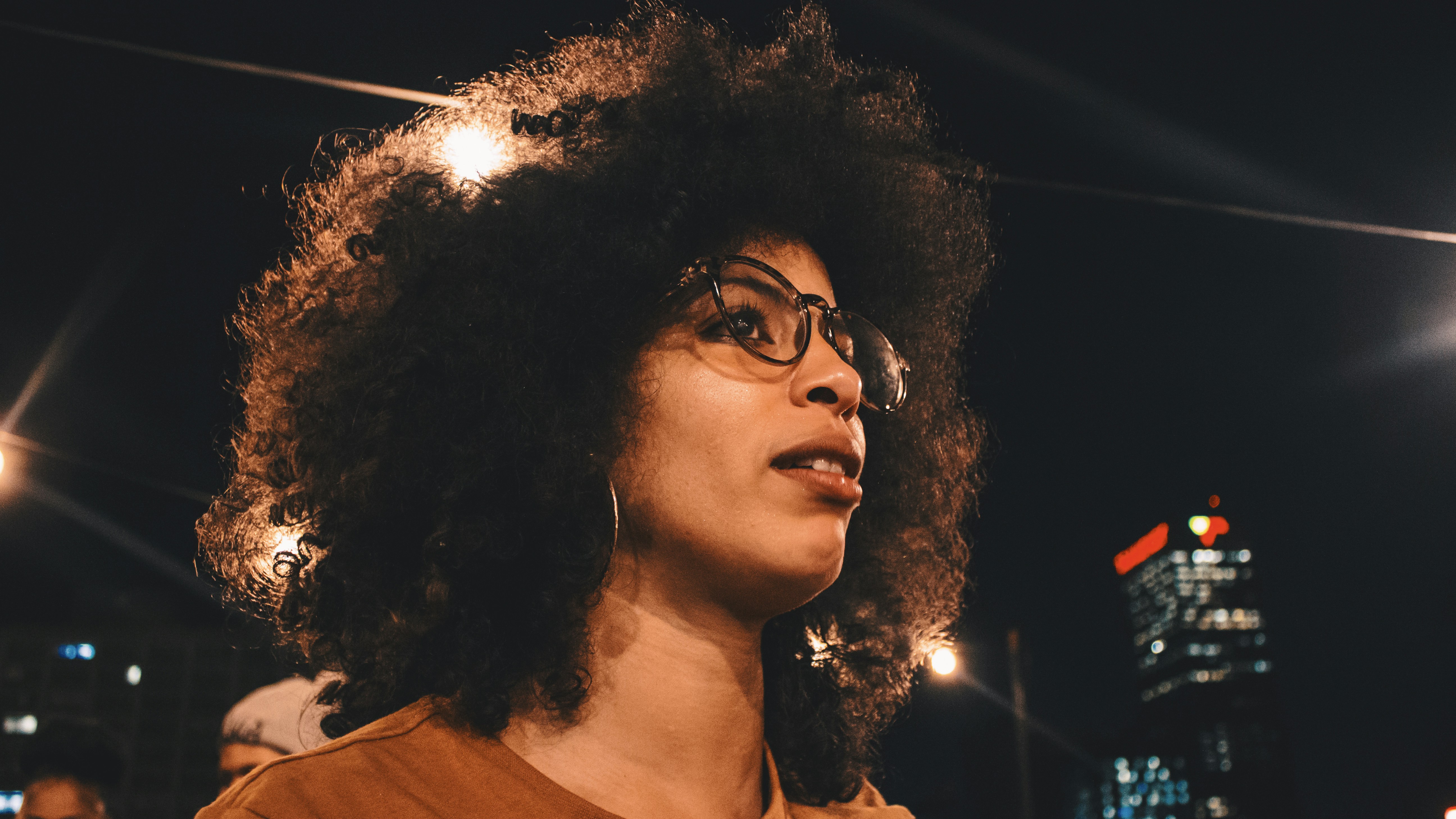 Woman with curly hair and glasses gazes thoughtfully into the distance, illuminated by city lights at night.