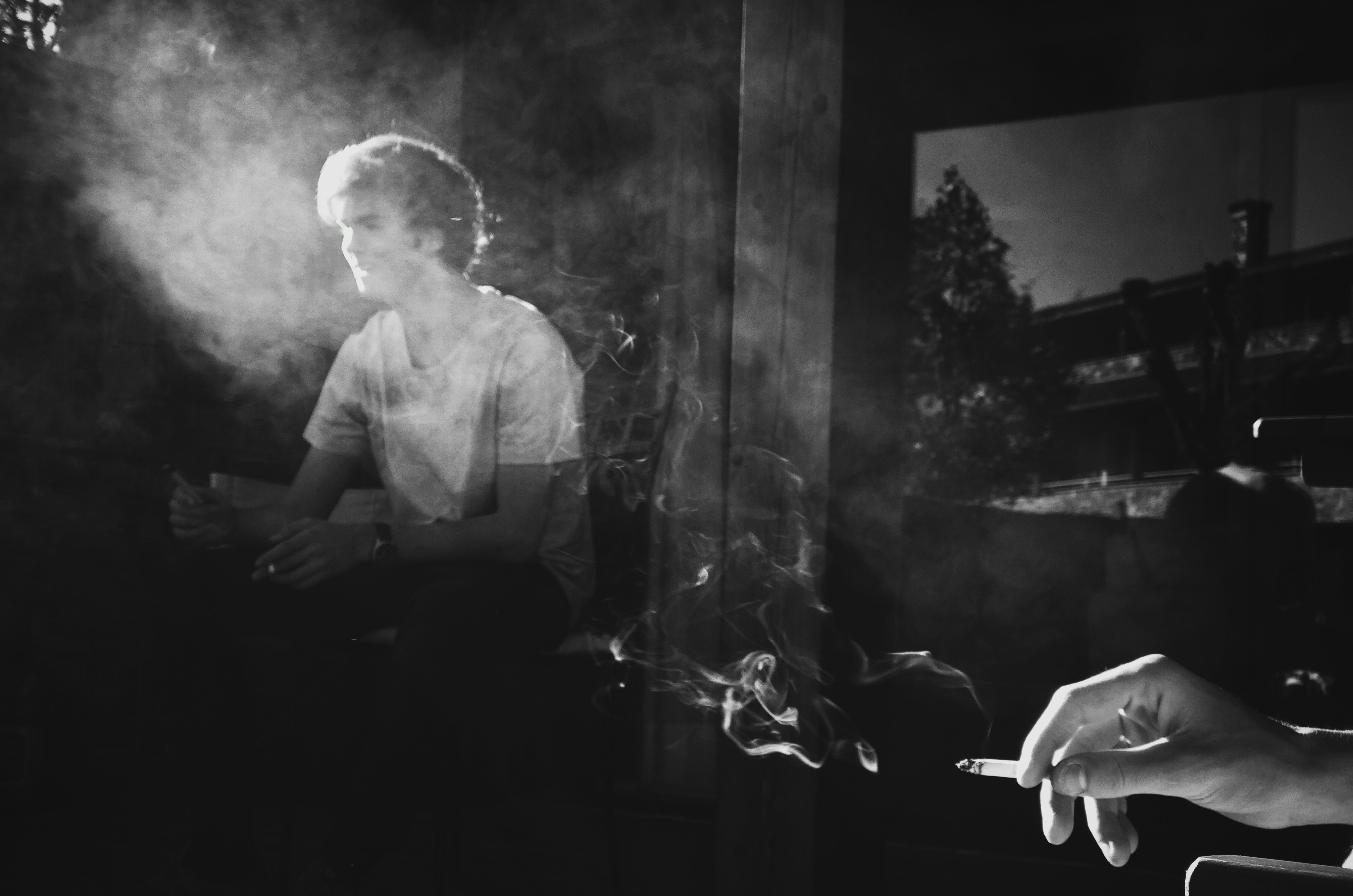 A contemplative young man sits in a dimly lit space, surrounded by swirling smoke, creating an intimate atmosphere. The contrast between light and shadow enhances the mood.