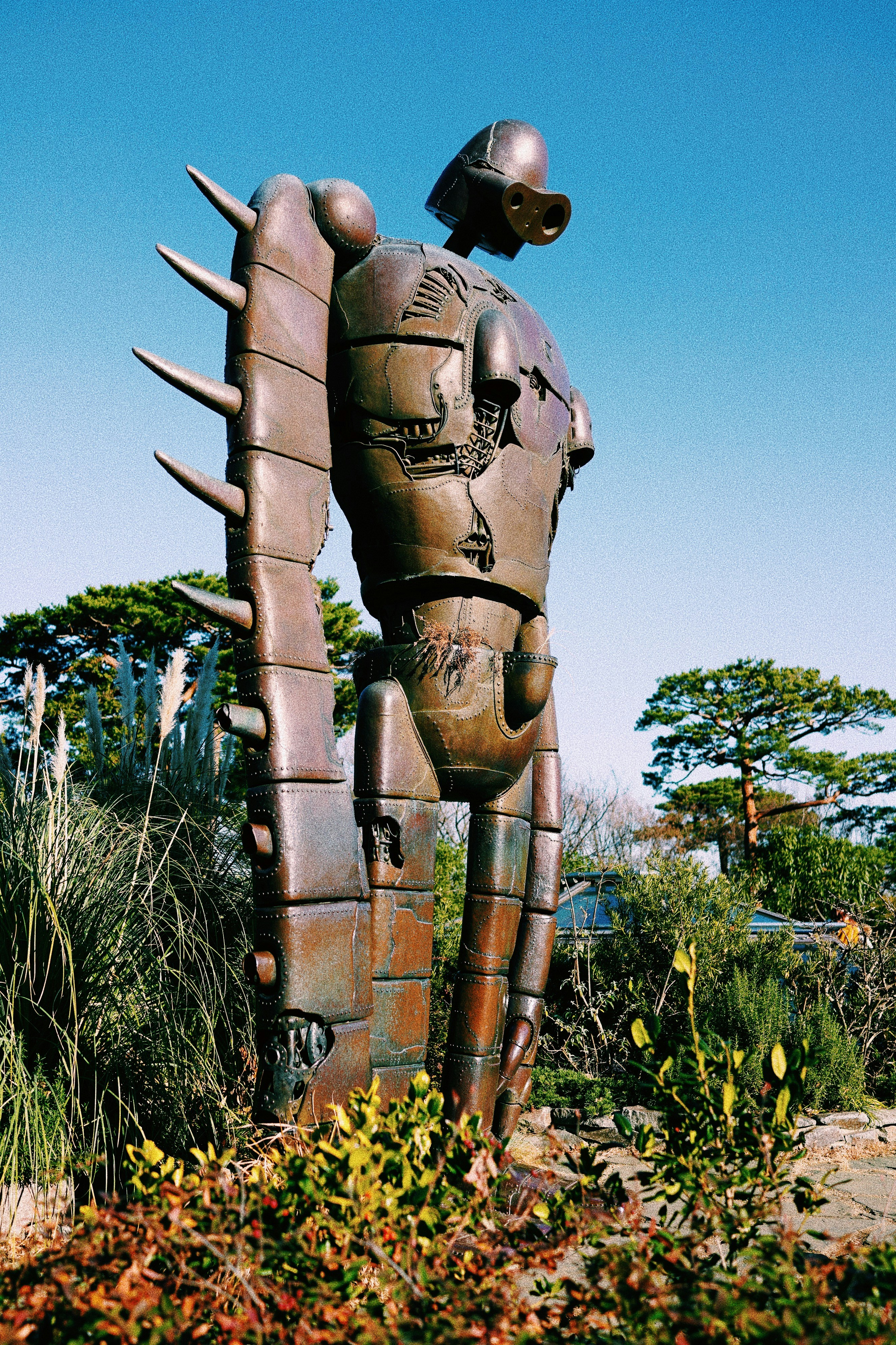 A towering bronze sculpture of a robot stands amidst vibrant greenery, blending art with the natural landscape. The intricate details of the statue highlight its craftsmanship.
