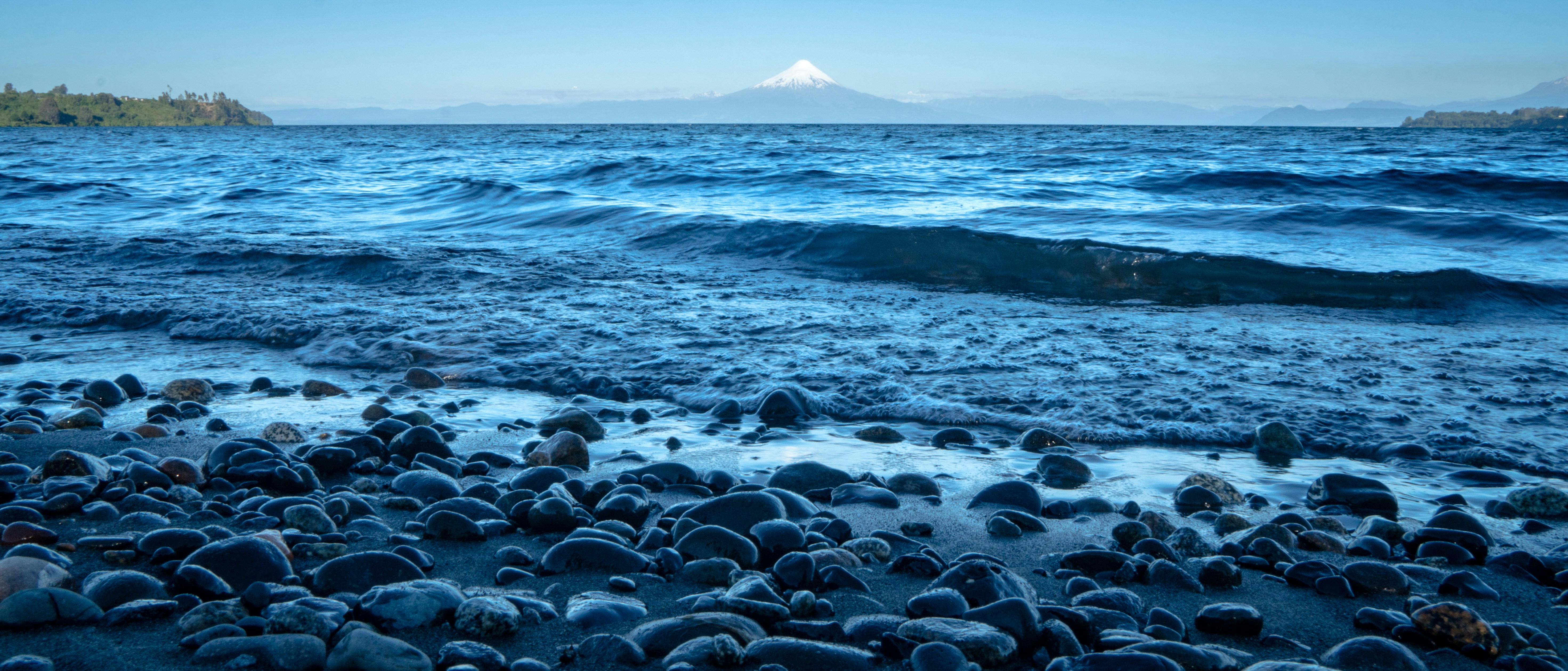 Pebble-Sea Horizon: Blue Tones and a Snow-Capped Sentinel