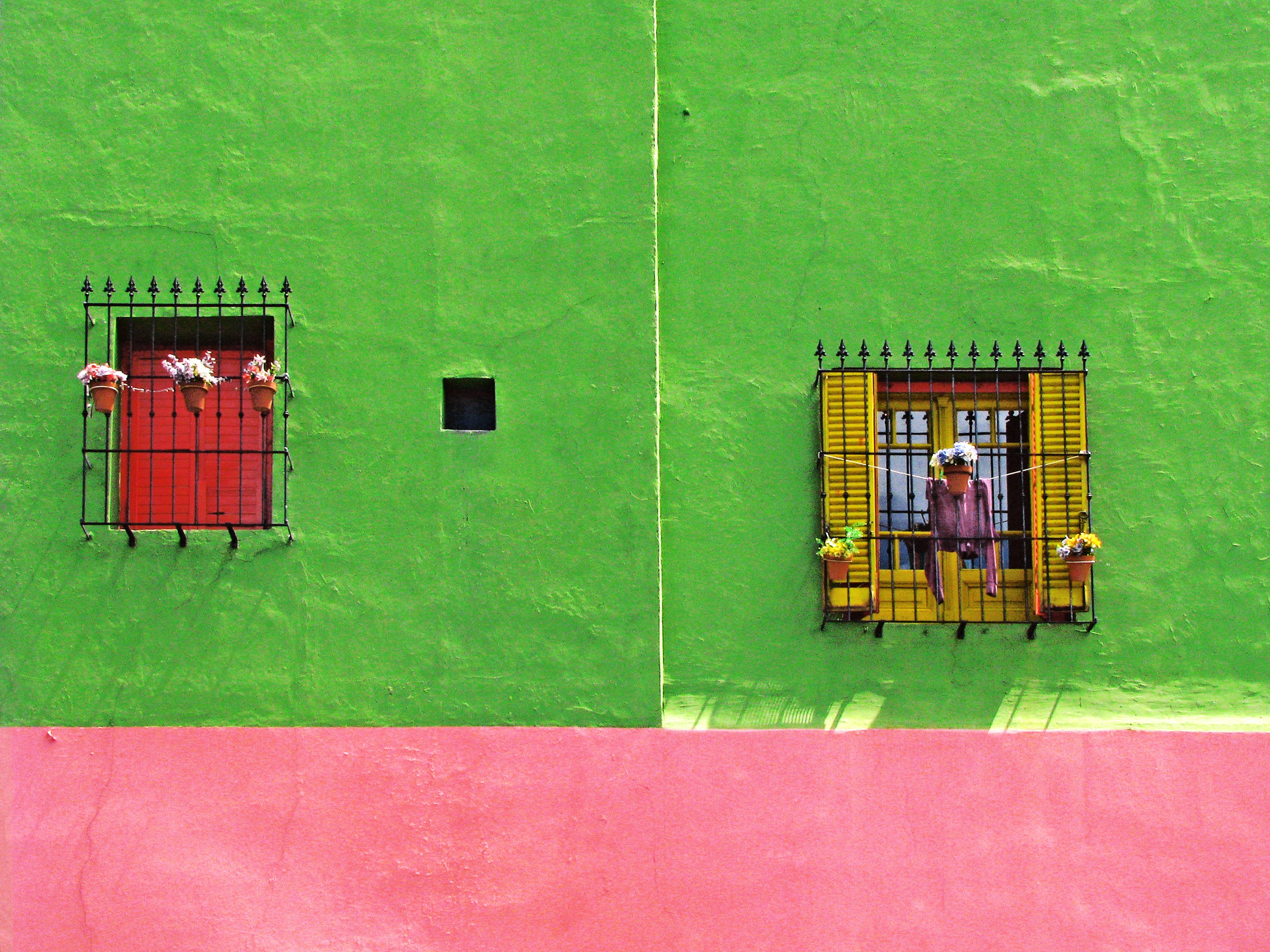 Two colorful windows on a vivid green wall, one with red accents and the other with yellow shutters.