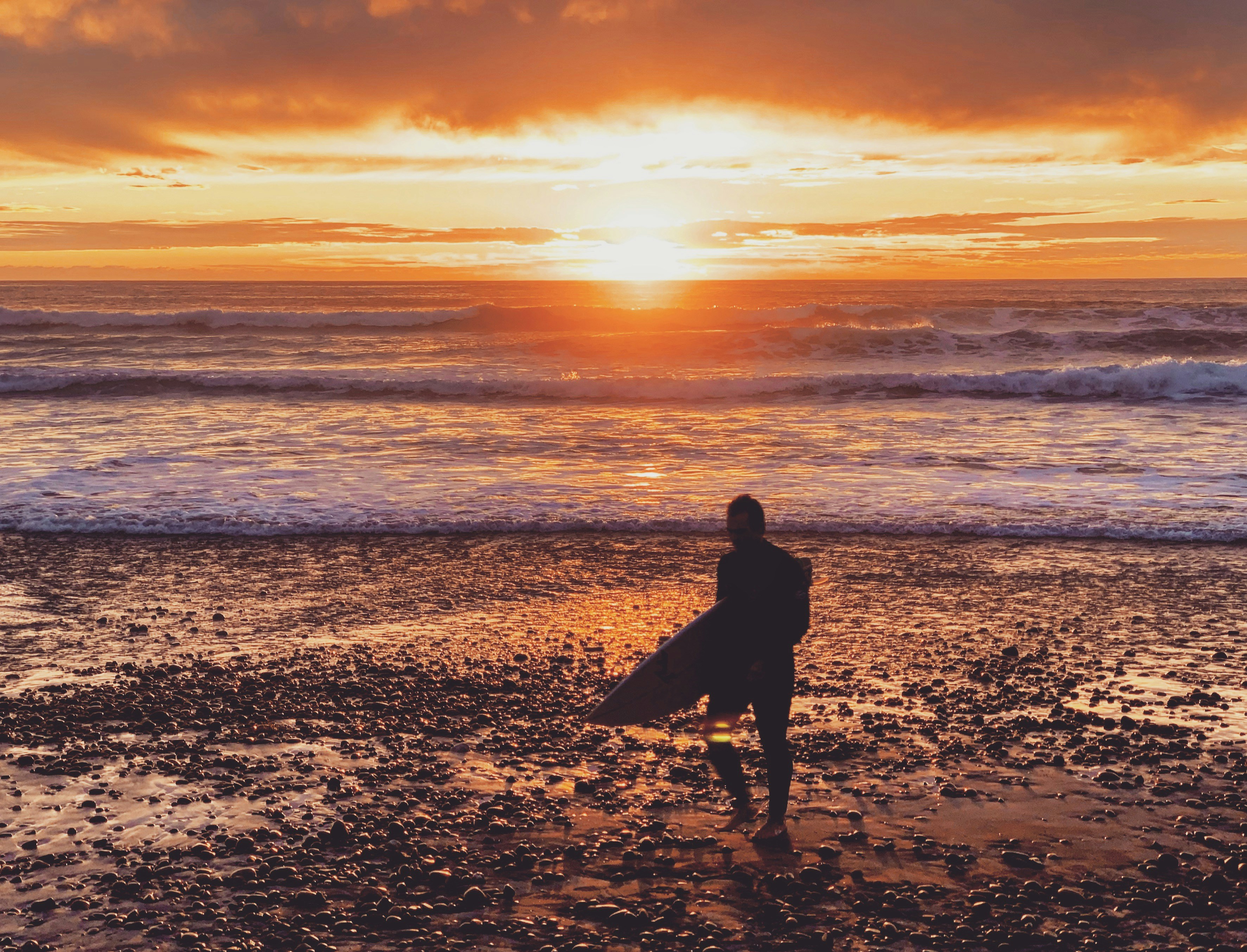 Surfer silhouetted against a vibrant sunset, walking along a rocky beach as waves gently lap at the shore.