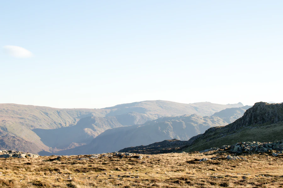 Dry grassy foreground leading to layered mountain ridges under a pale blue sky. A rocky slope rises on the right, adding depth to the wide landscape.