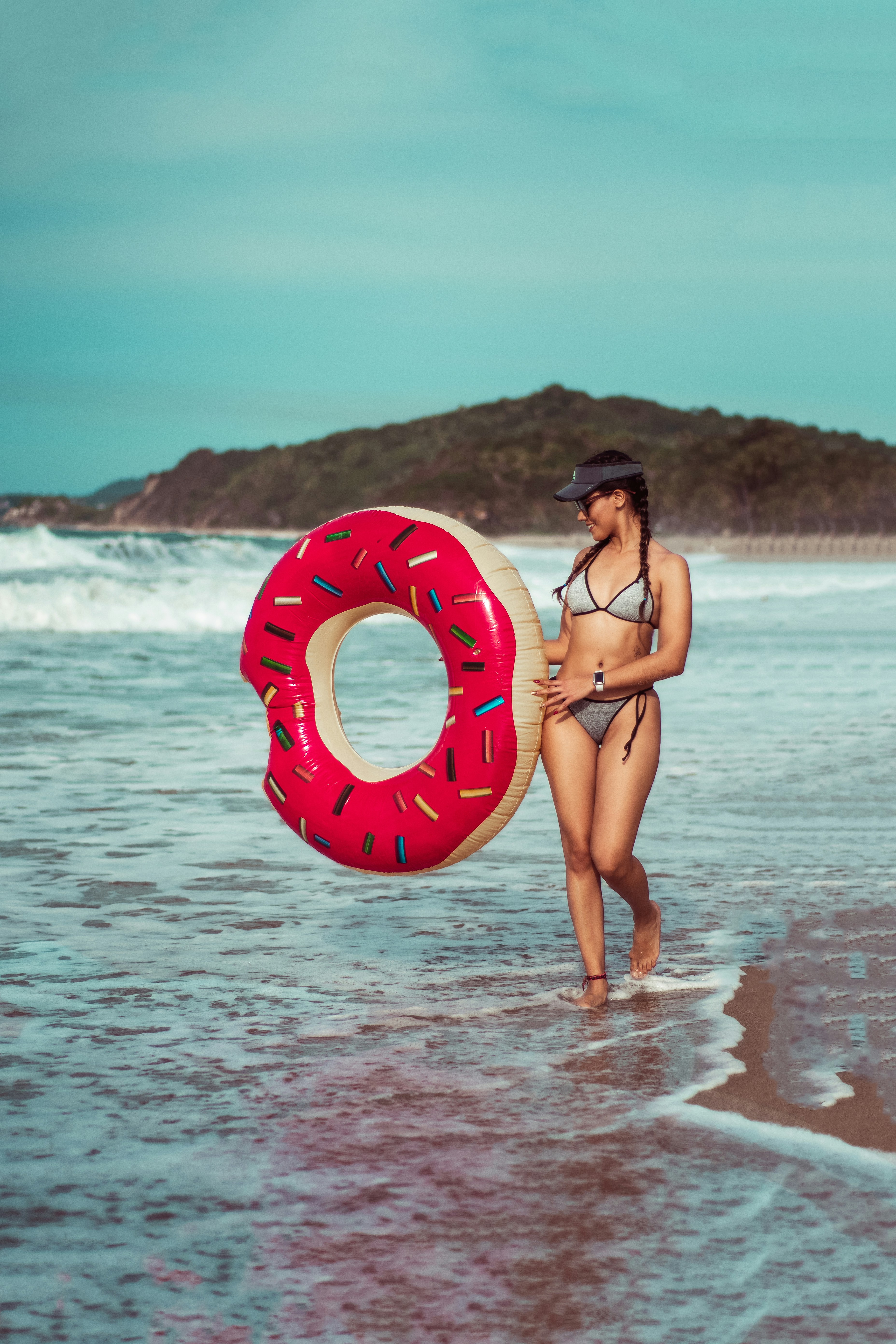 Woman walking along the shoreline holding a vibrant donut-shaped float, with gentle waves and a lush background.