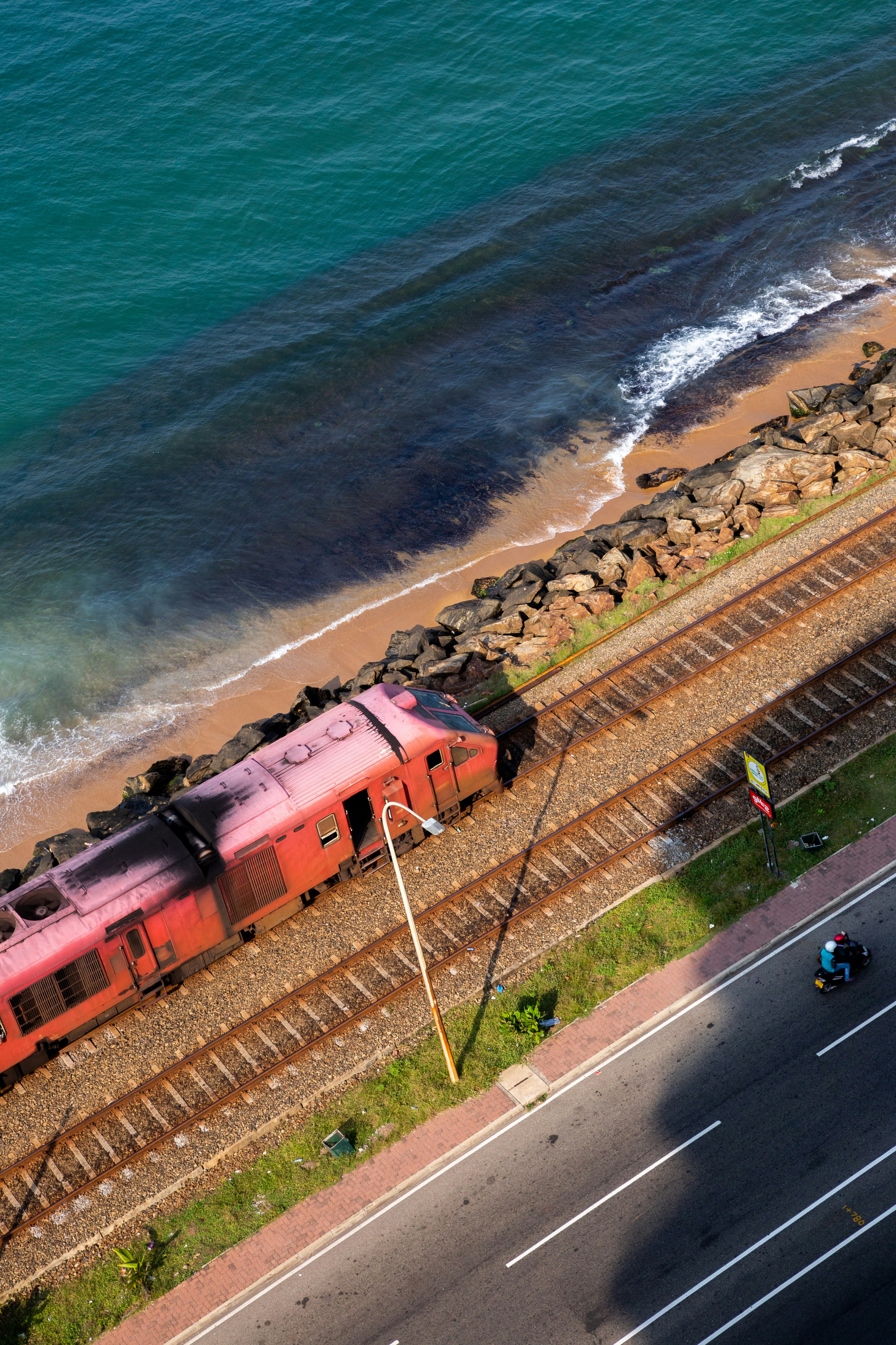 Abandoned pink train resting alongside a tranquil beach, with waves lapping at the shore and a road nearby.