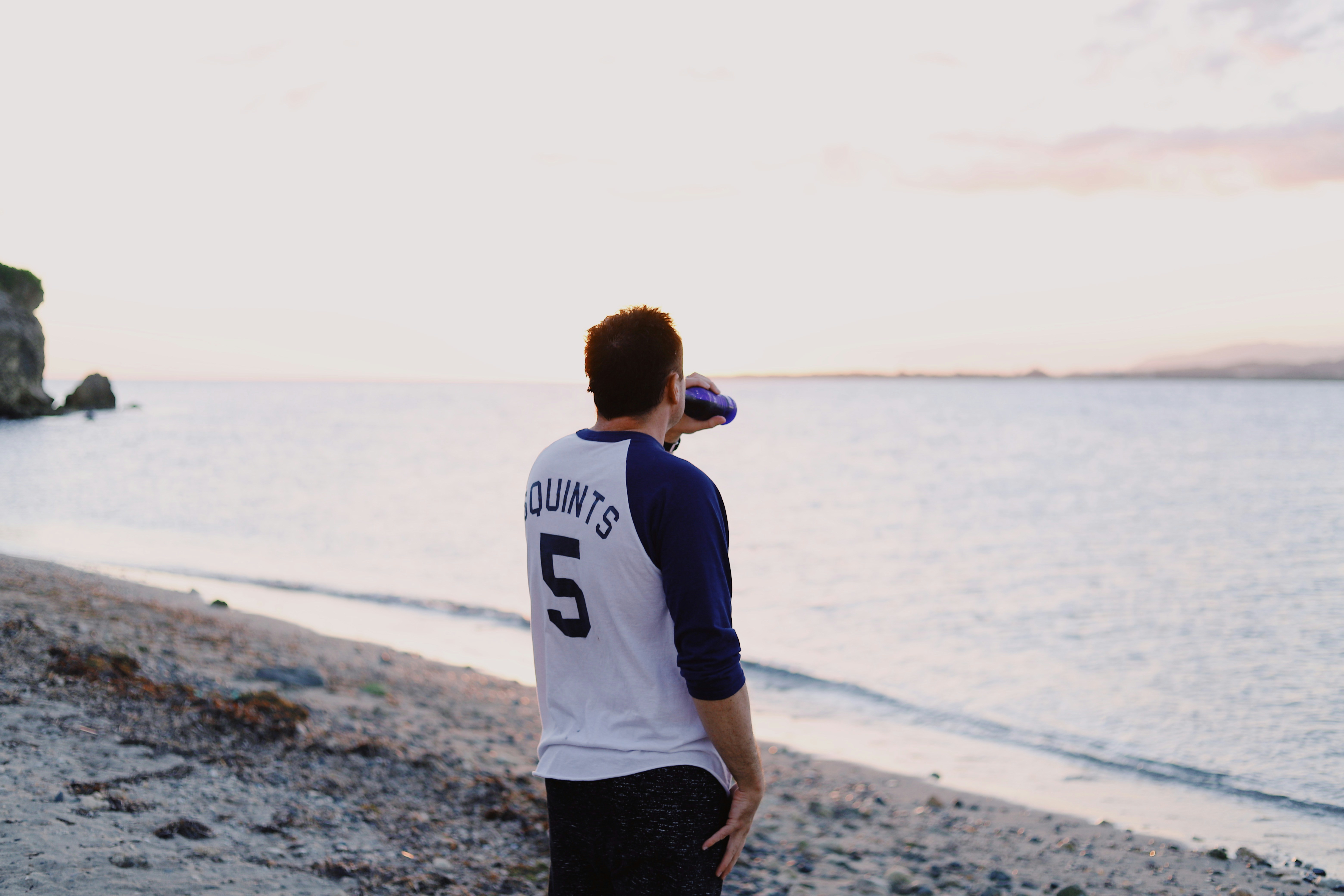 A man in a baseball jersey gazes at the tranquil sea during sunset, holding a drink. The serene beachscape enhances the contemplative mood.