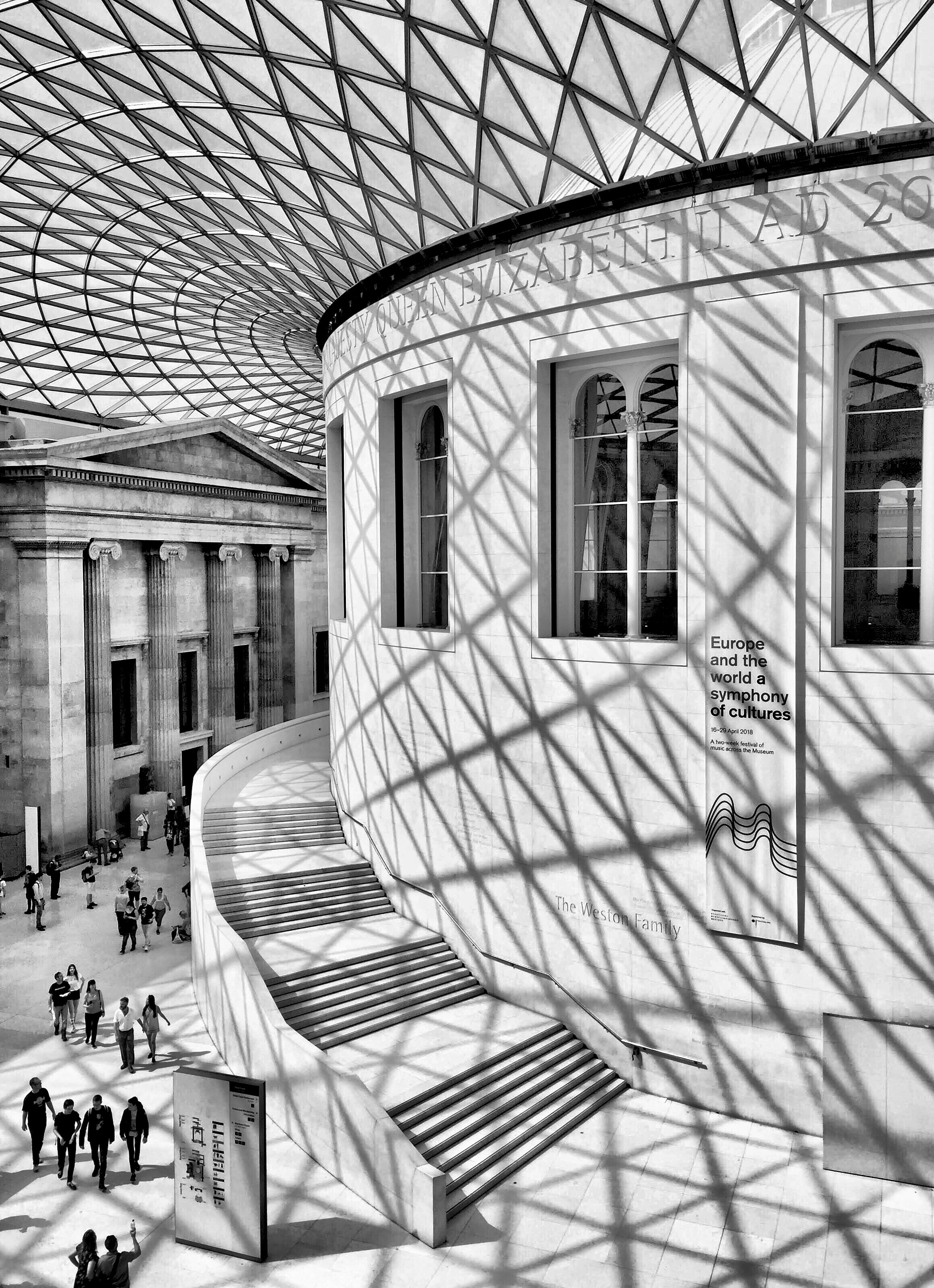 Intricate shadows cast by a glass ceiling create a dynamic interplay of light and architecture in a bustling museum space.