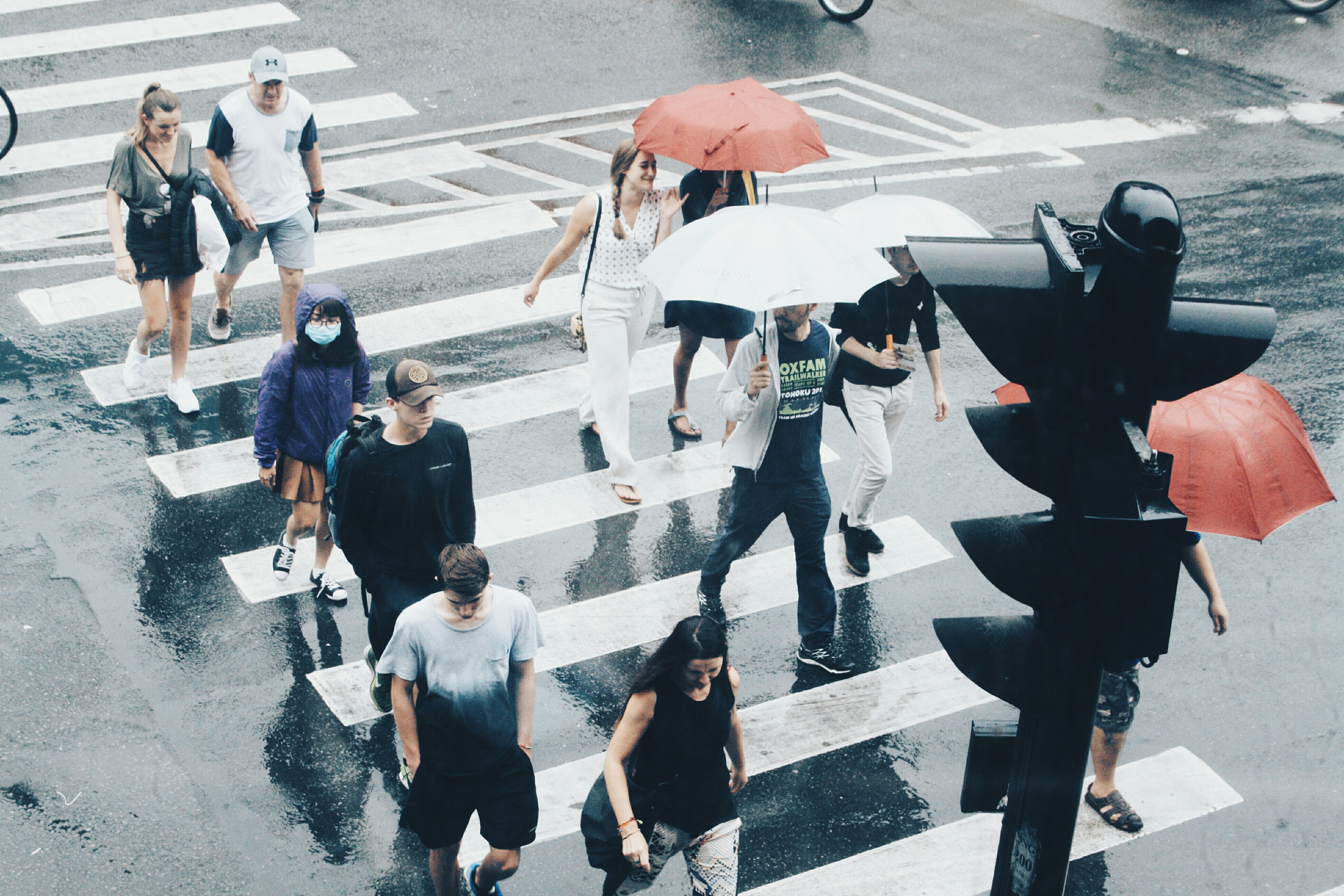 Rainy urban crosswalk photographed from above, pedestrians carry colorful umbrellas as rain slicks the pavement.