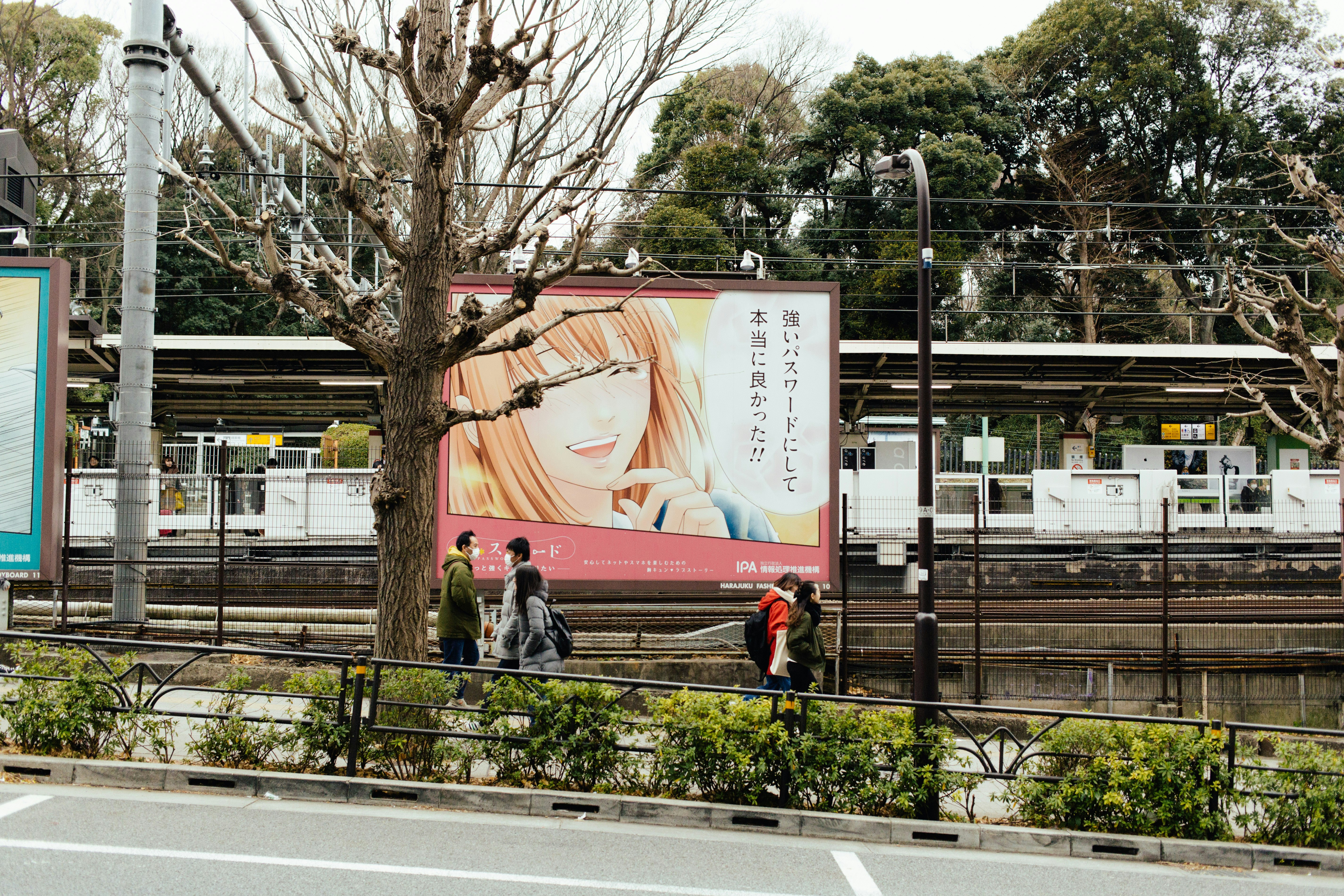 A vibrant advertisement featuring a smiling character stands prominently at a train station, while pedestrians stroll by amidst trees and urban greenery.