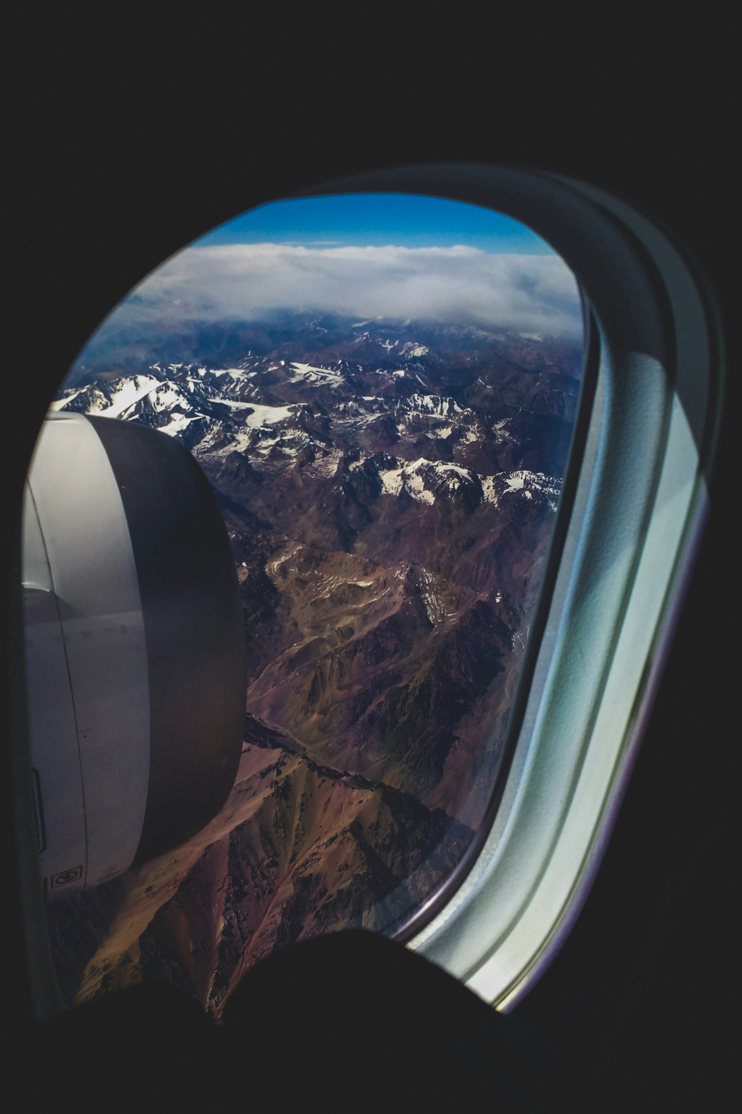 View of snow-capped mountains from an airplane window, framed by the aircraft's wing and engine. A serene sky adds depth to the landscape.
