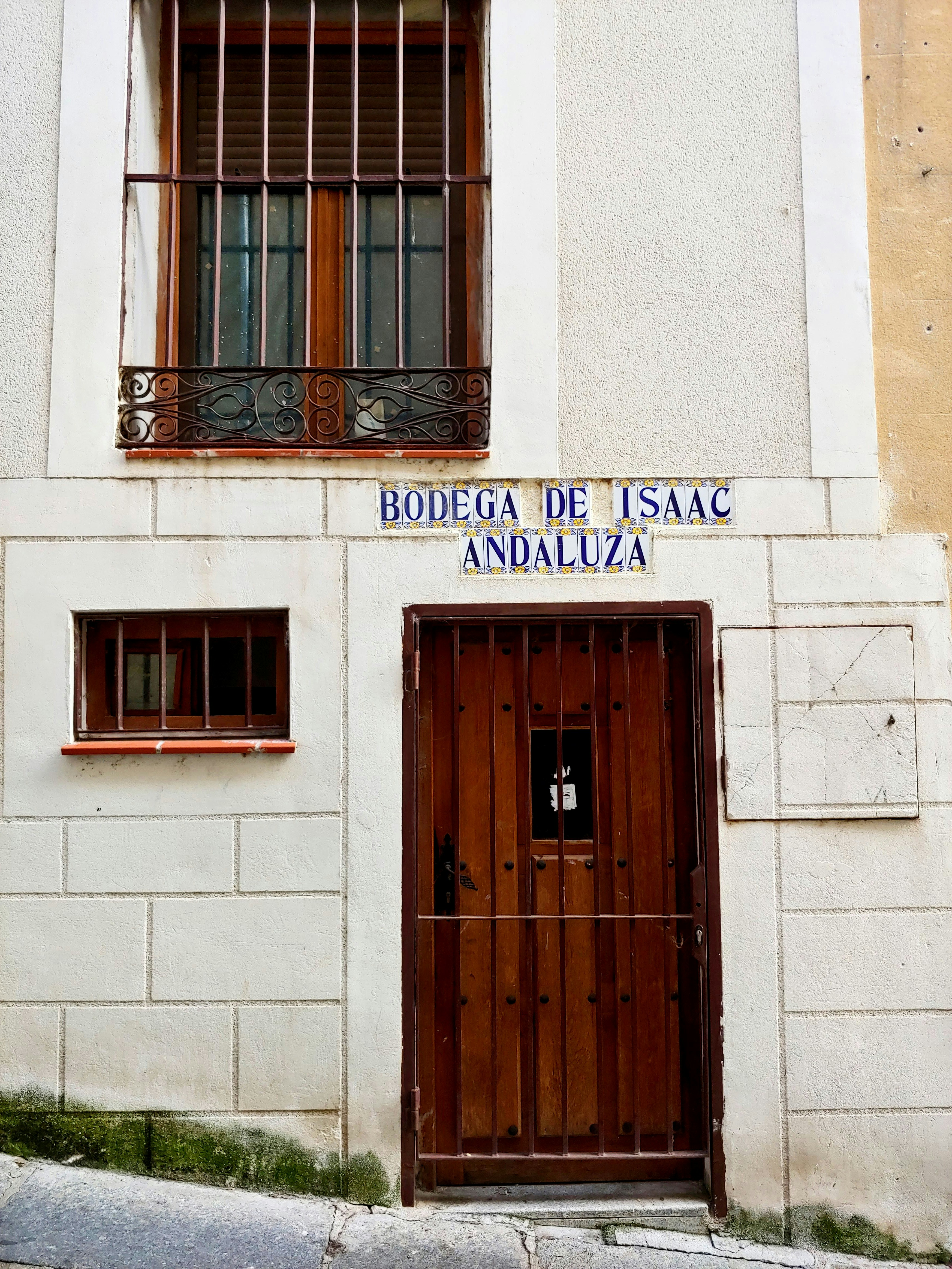 Wooden door with metal bars beneath a sign reading 'BODEGA DE ISAAC ANDALUZA' on a light stone facade; upper window with iron railing sits above. An architectural doorway scene highlighting Spanish signage and rustic detailing.