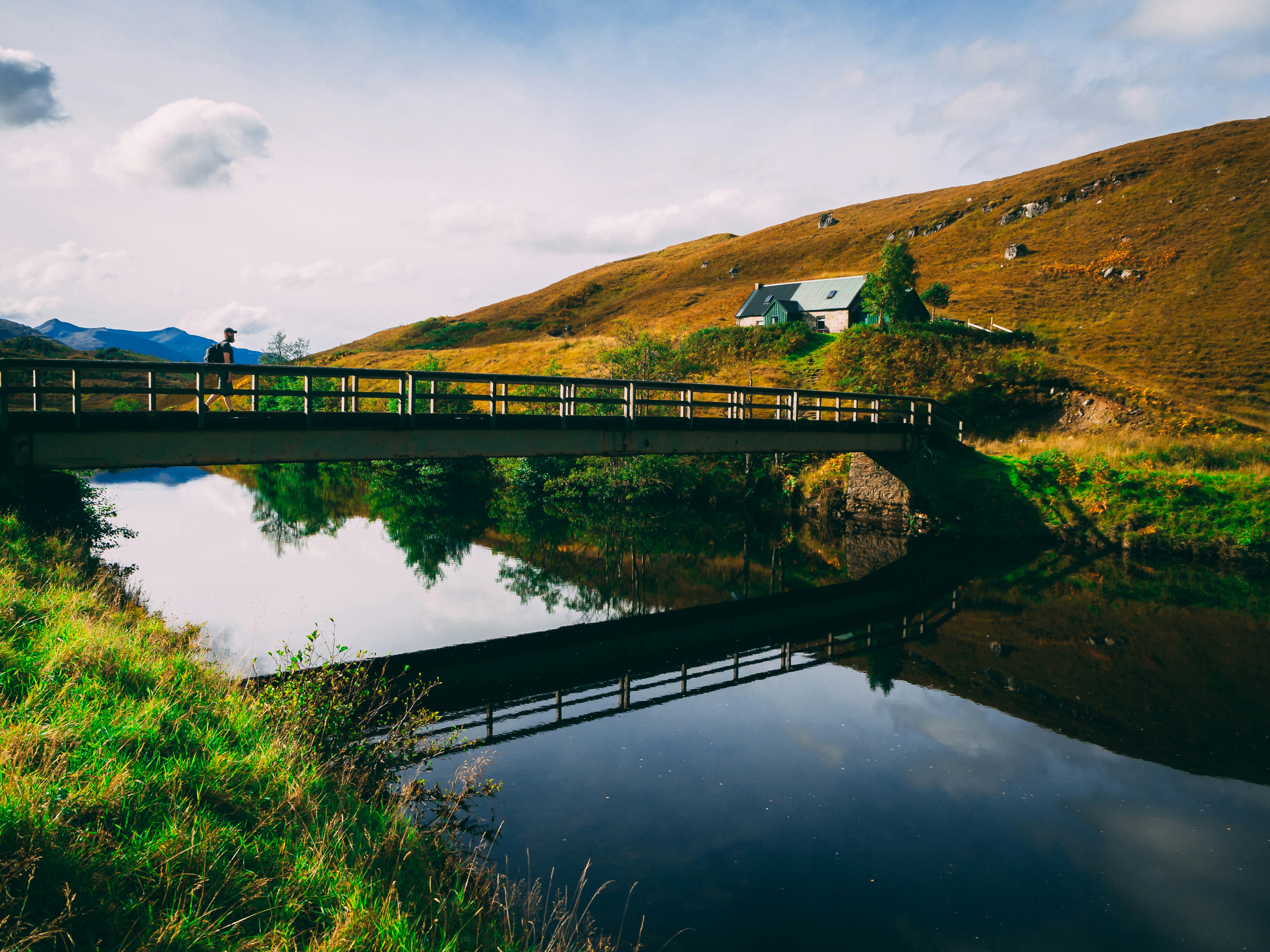 A wooden bridge spans a calm river, surrounded by rolling hills and a quaint house. The scene reflects a peaceful rural landscape.