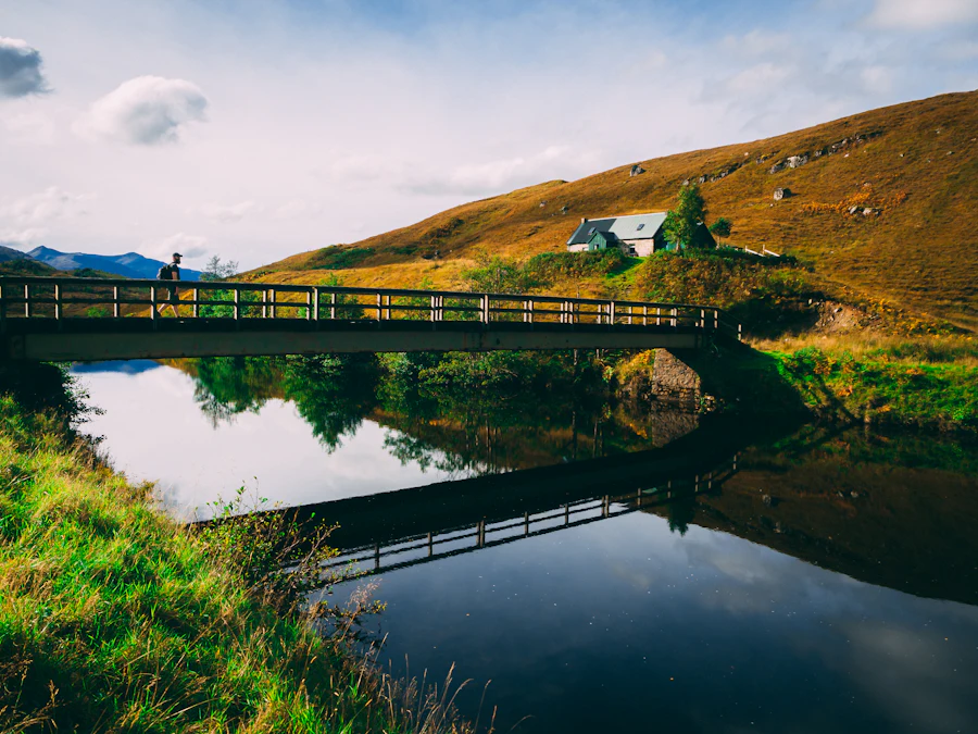 A small bridge crosses a calm stream reflecting the sky and hillside, with a lone house on the slope beyond. Golden grass and distant mountains frame the rural landscape.