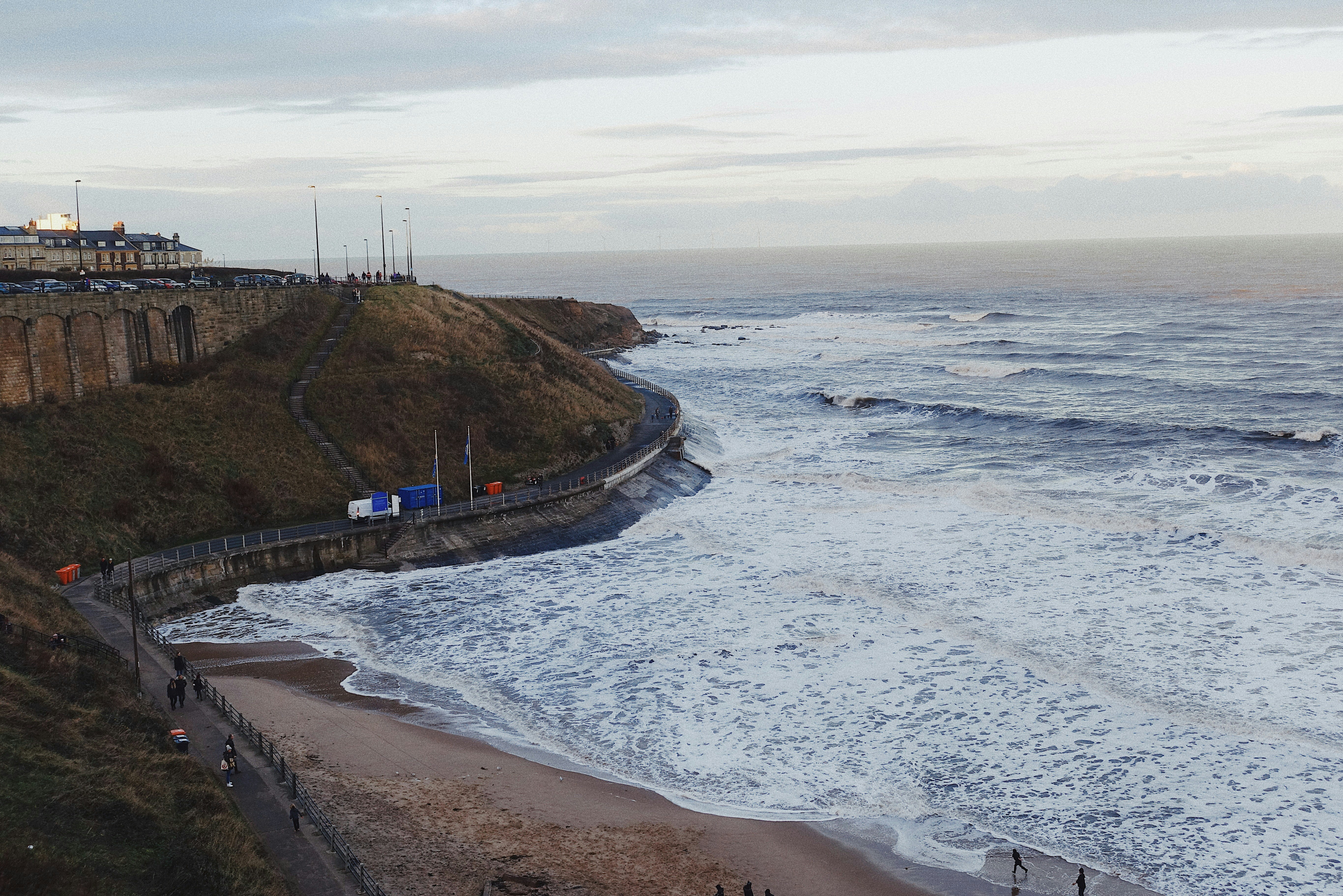 Waves crash against the shore, with a winding path and people walking along the beach at dusk. The coastline is framed by cliffs and the remnants of a seaside town.
