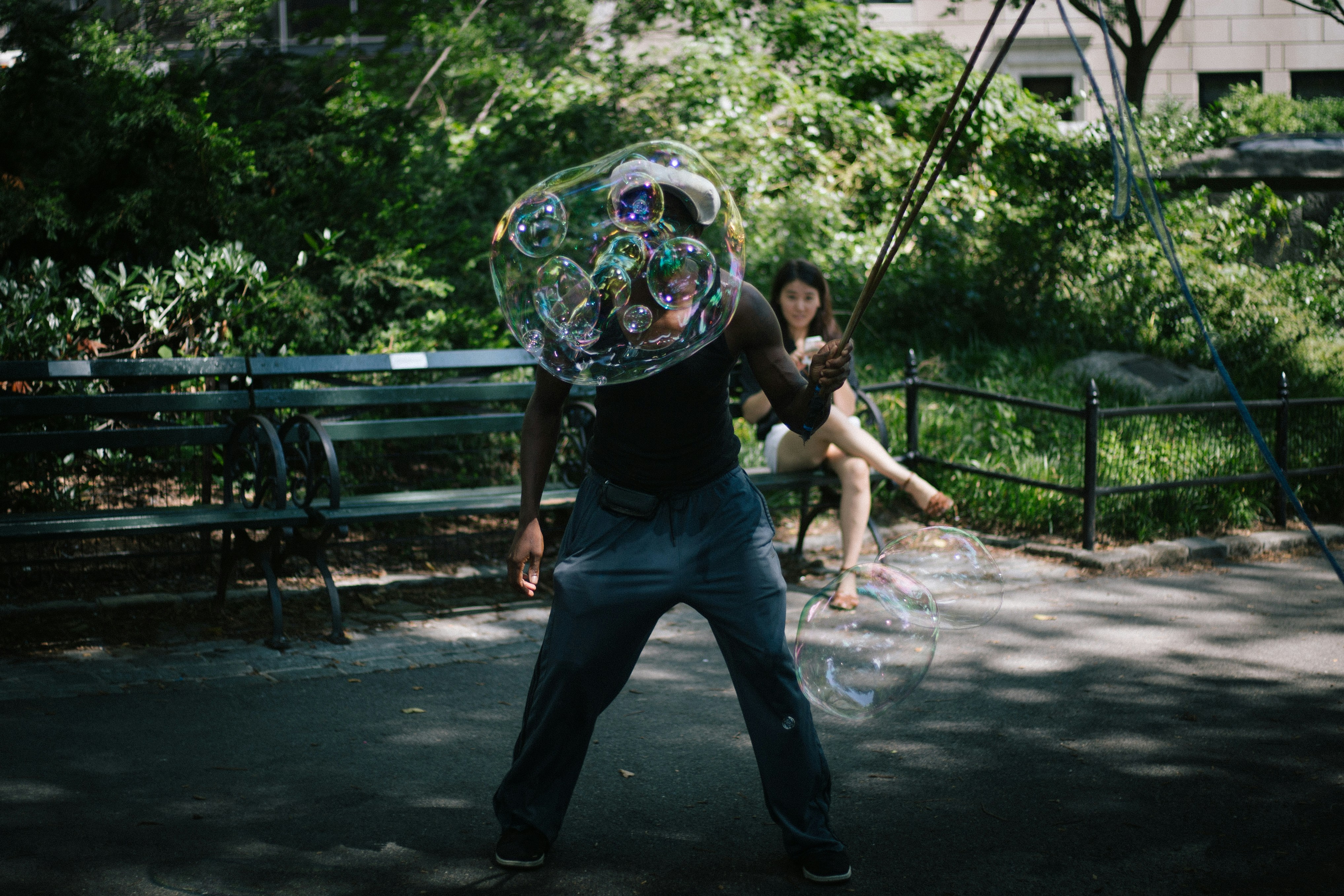Man creating a large soap bubble in a park, with a woman seated on a bench in the background.