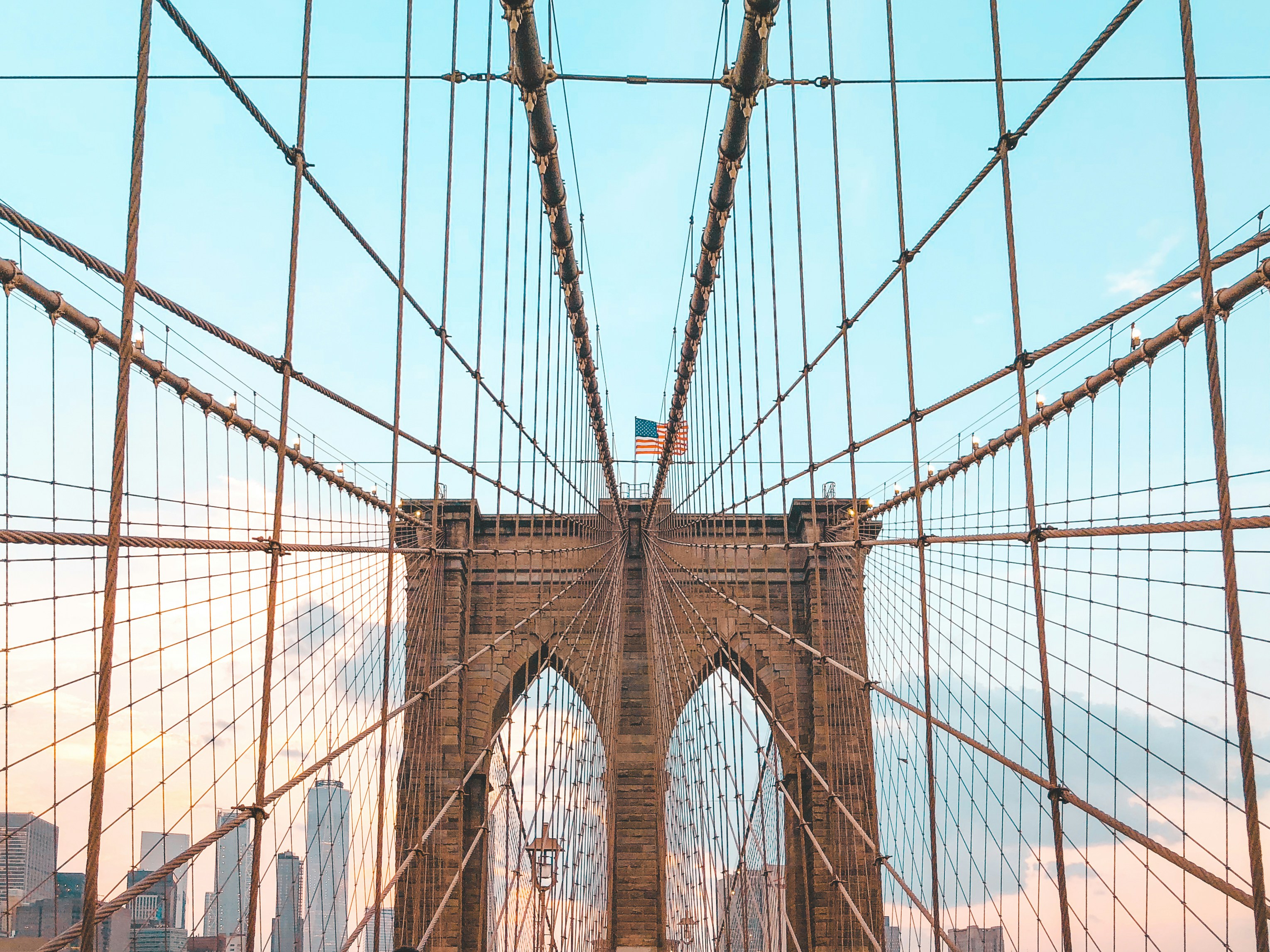 Brooklyn Bridge's intricate cable structure converging towards the American flag against a pastel sky.