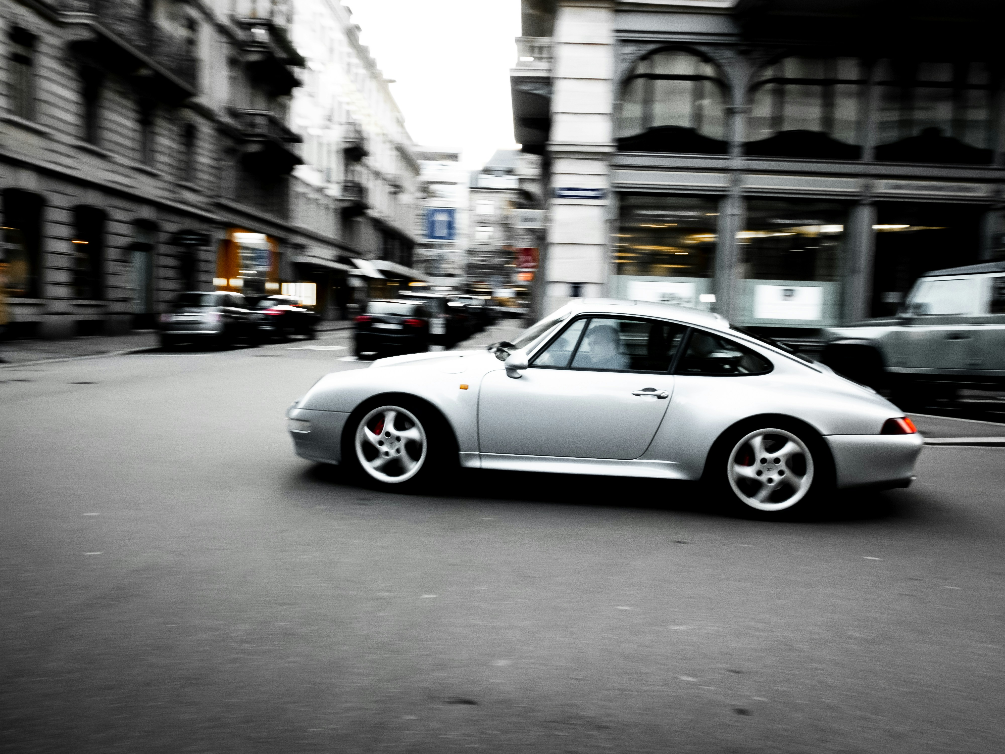 A silver sports car navigating a city street, showcasing its sleek design against a backdrop of blurred buildings and traffic. The dynamic motion conveys speed and sophistication.