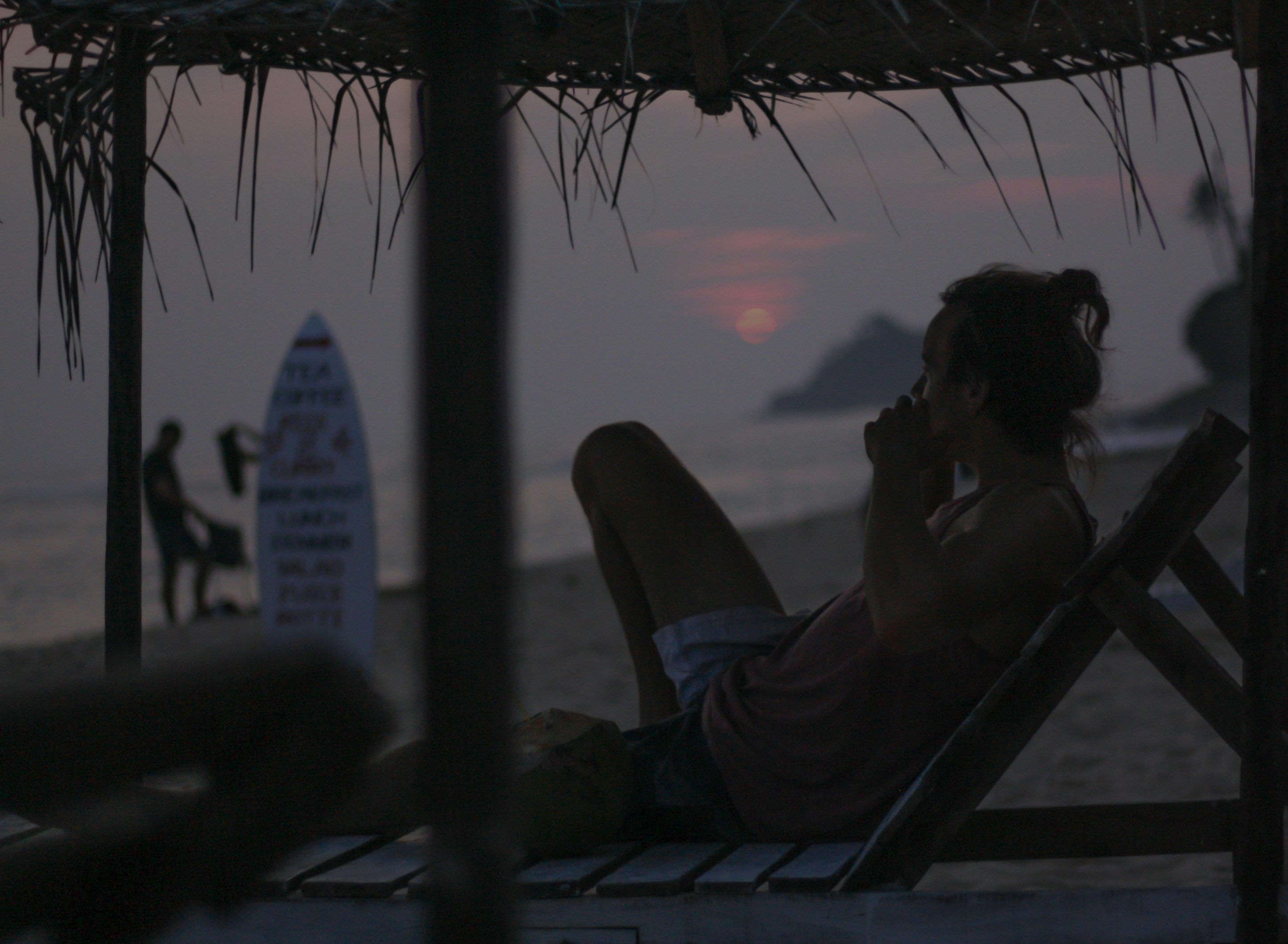Silhouette of a person lounging on a beachside wooden deck, sipping a drink as the sun sets in the background, creating a tranquil atmosphere.